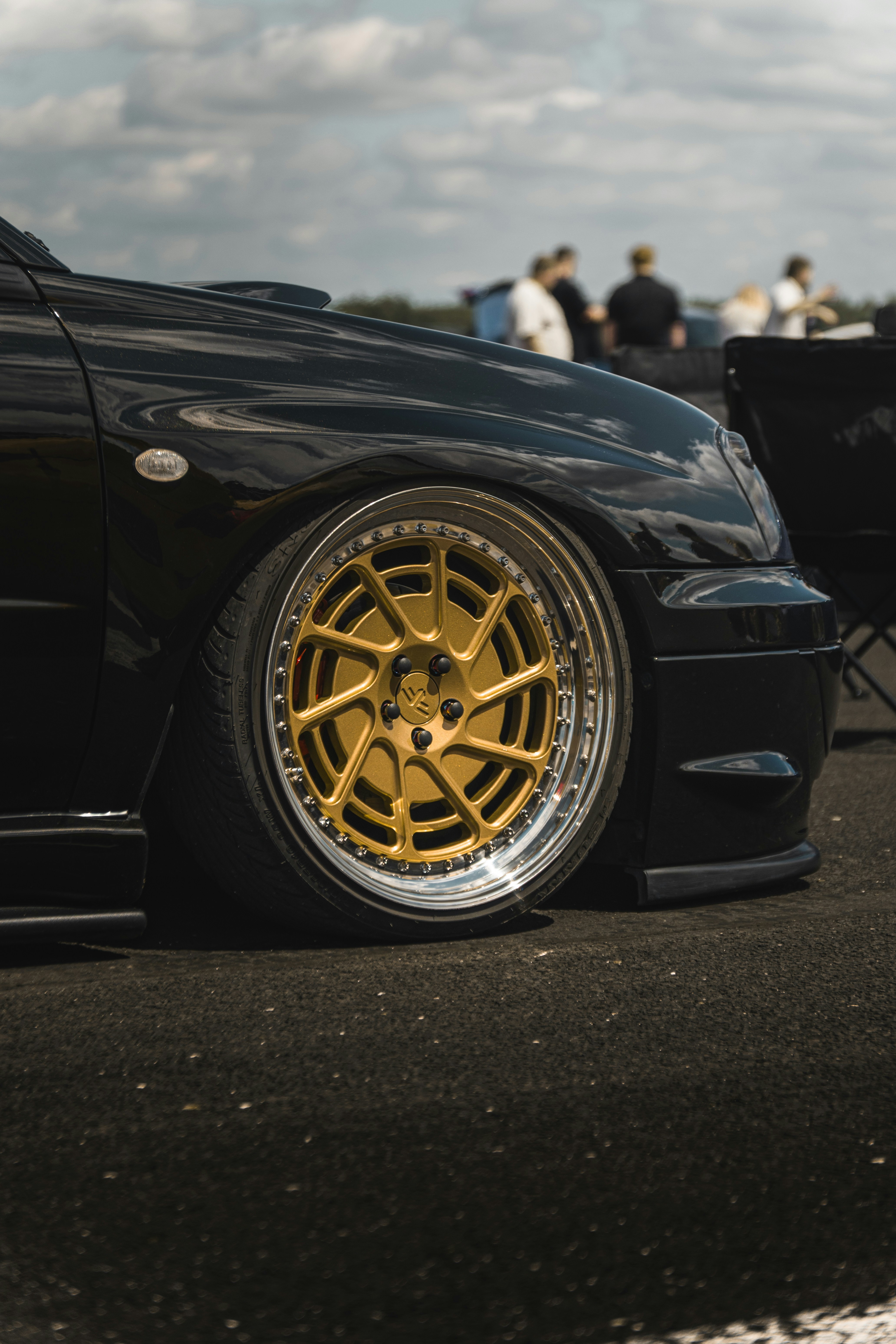 Low-angle view of a black car with a gold multi-spoke wheel, set against a blurred crowd at an outdoor event. Emphasis on wheel detail and glossy body work.