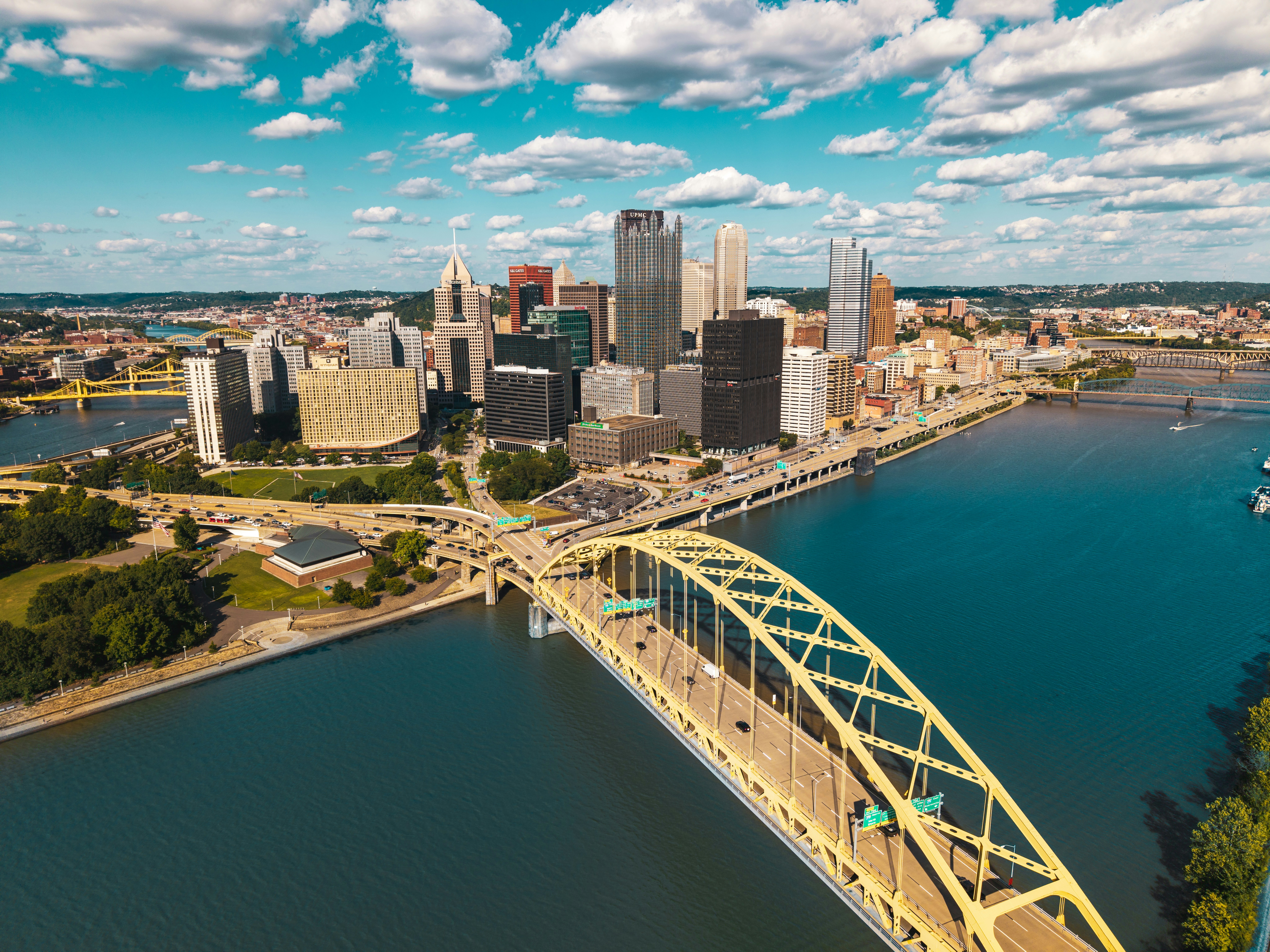 Aerial view of a city skyline with a prominent yellow arch bridge spanning a blue river, surrounding high-rise buildings, and a clear sky.