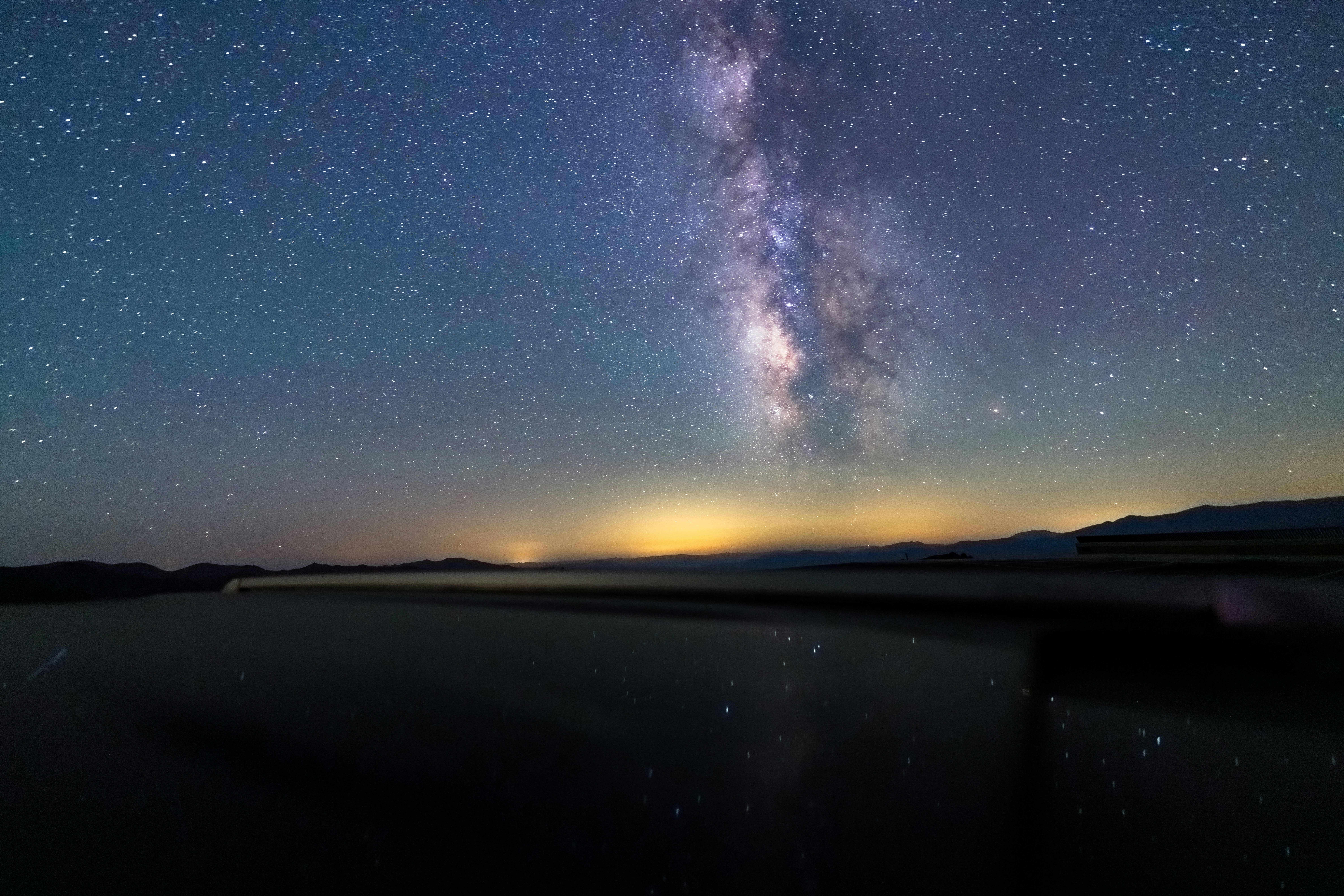 Star-filled sky with the Milky Way stretching upward, reflecting softly on a dark, tranquil body of water near a subtle horizon glow.