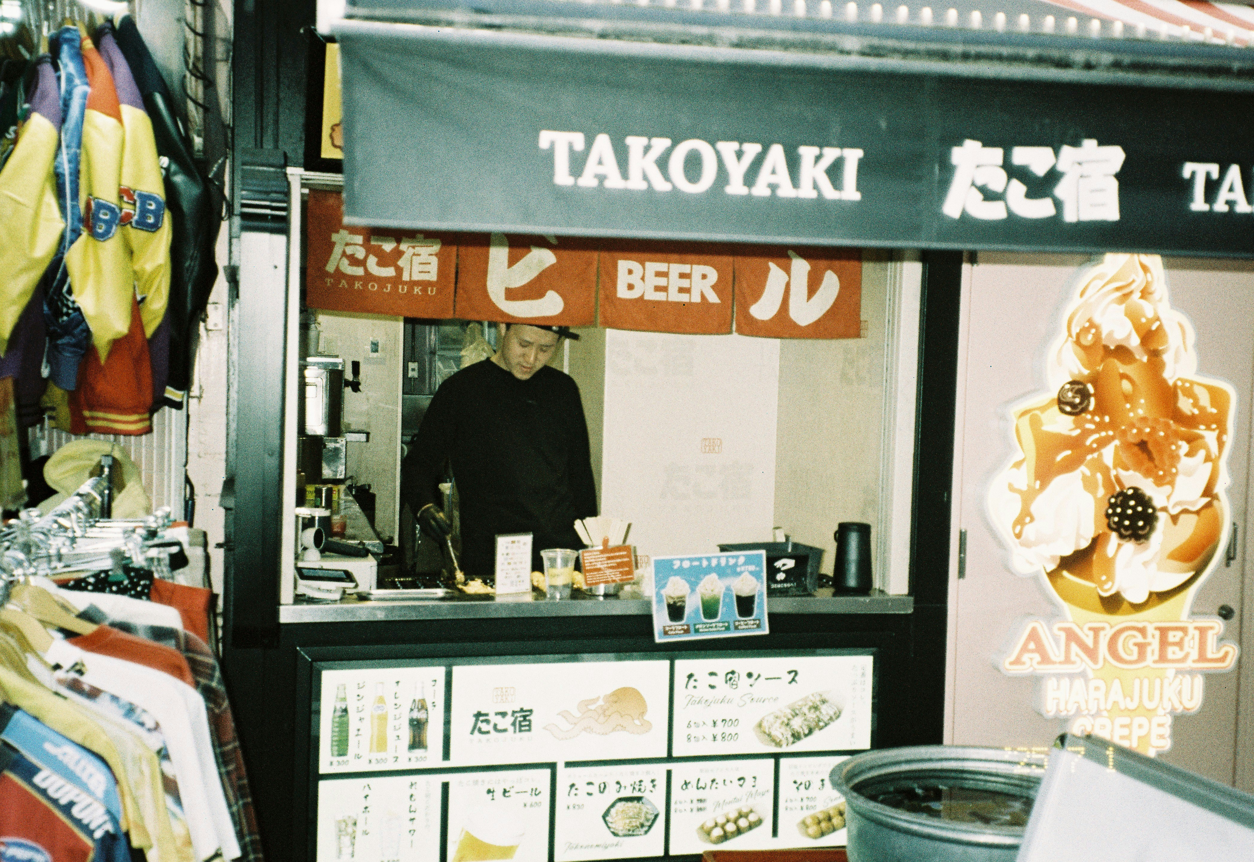 Small takoyaki stall with a vendor behind the counter; a bright poster featuring an ice-cream-like dessert and clothing booths frame the scene.