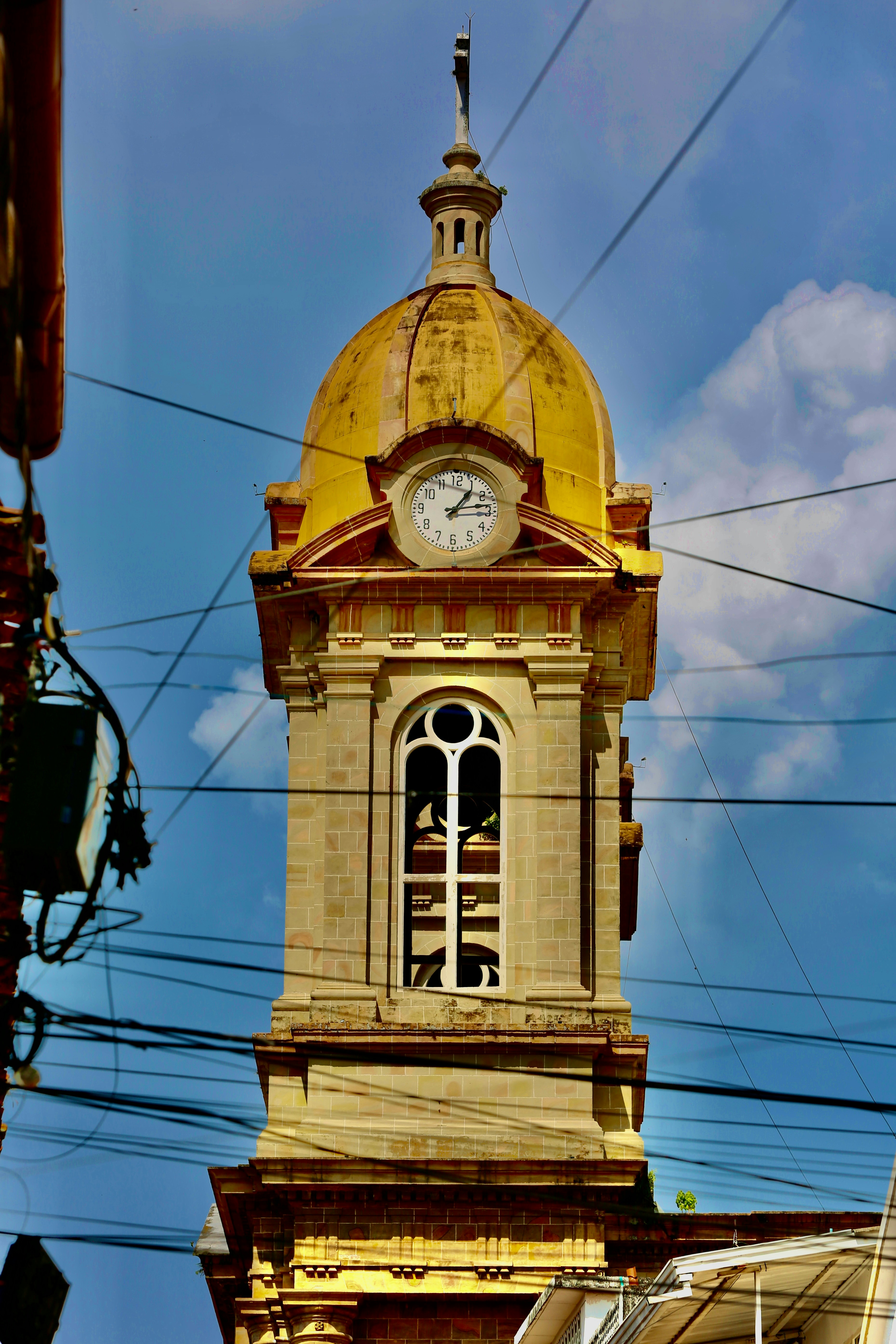 Clock-topped tower bathed in golden light against a blue sky, with overhead wires crisscrossing the foreground and a hint of urban architecture at the base. A city landmark captured with architectural detail.