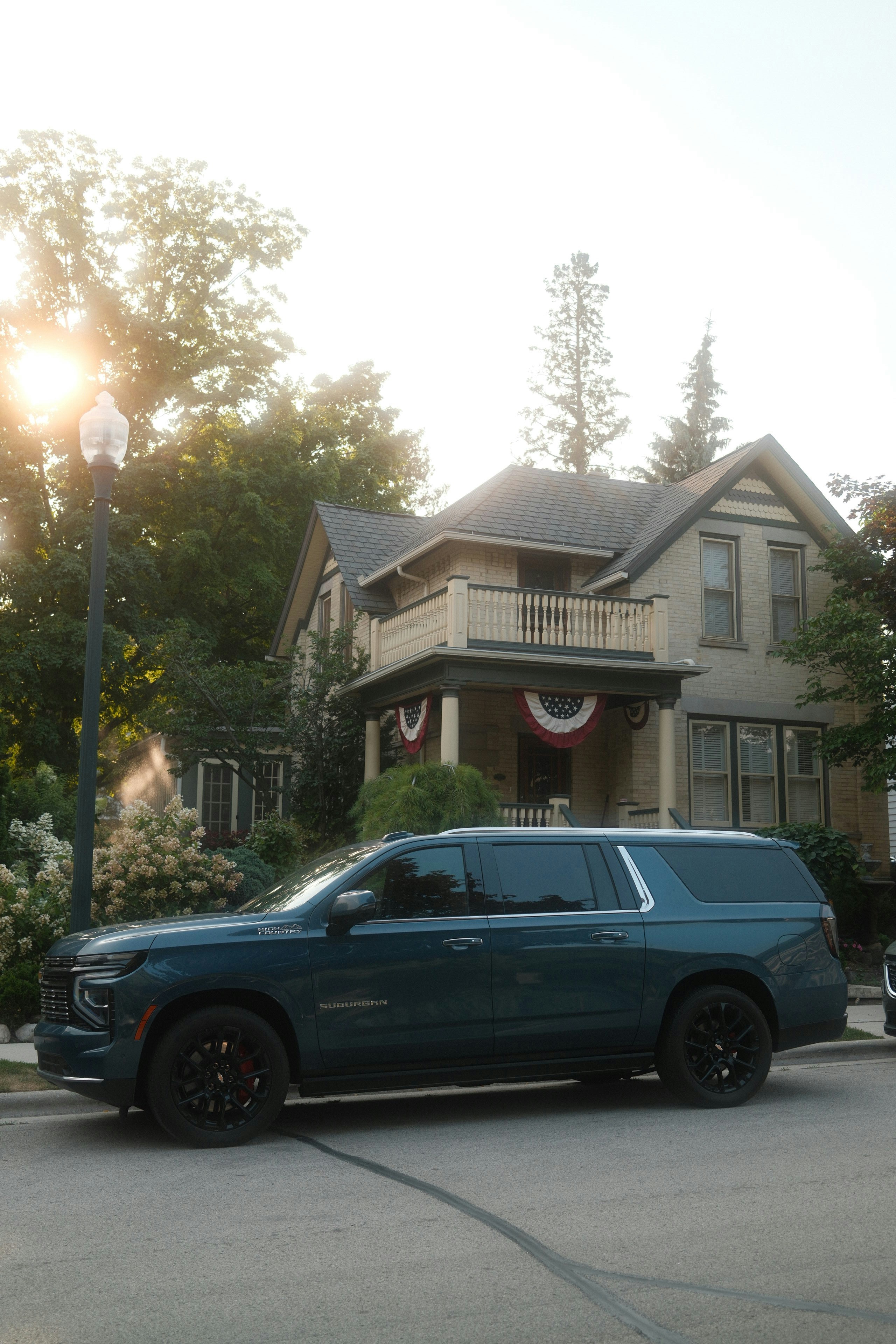 A blue SUV parked in front of a two-story brick house with bunting, under a warm sunset glow. The scene includes a lamppost to the left and surrounding greenery.