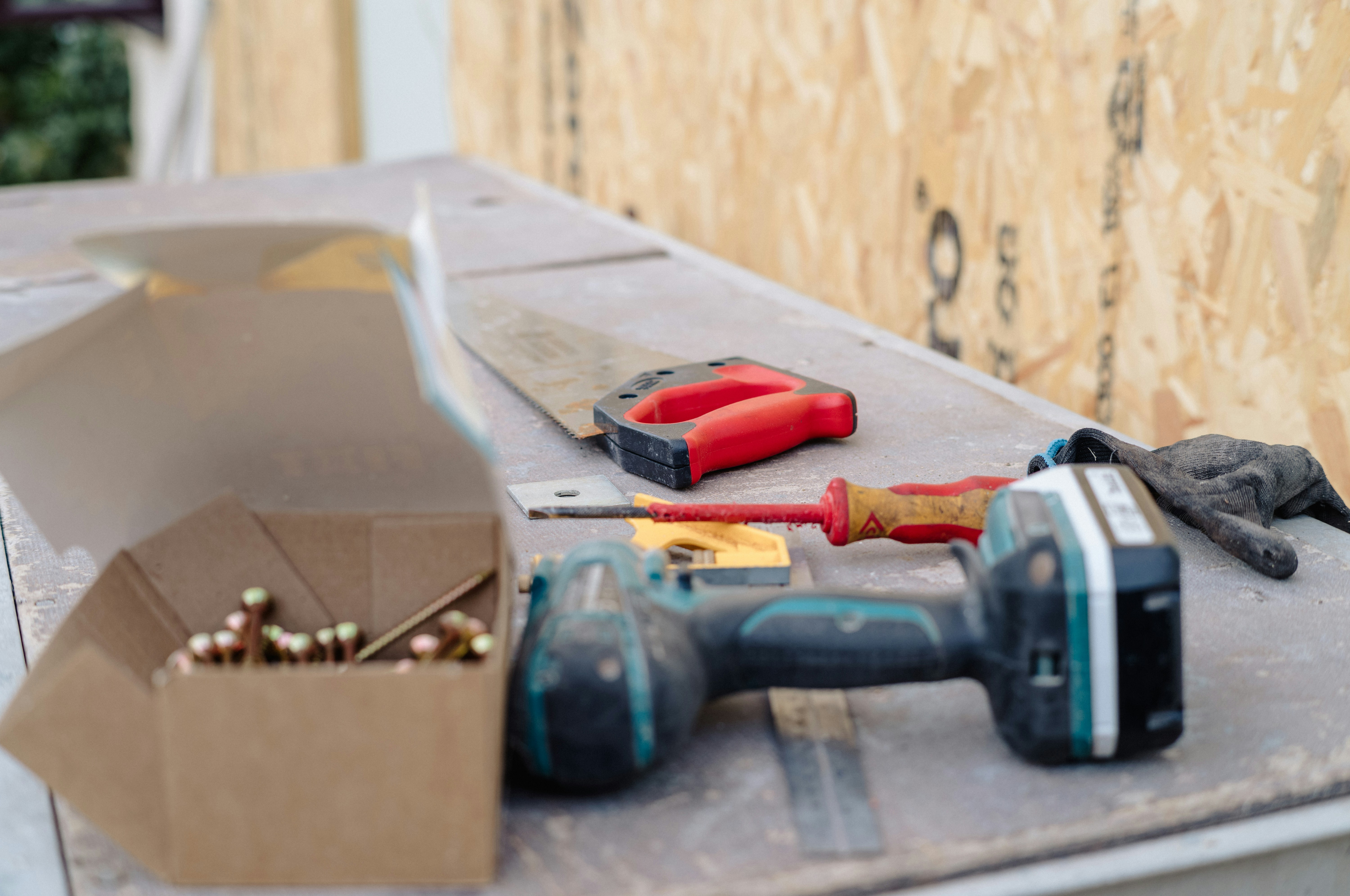 Tools and a box of spent cartridges sit on a dusty worktable at a construction site, with an unfinished plywood wall in the background.