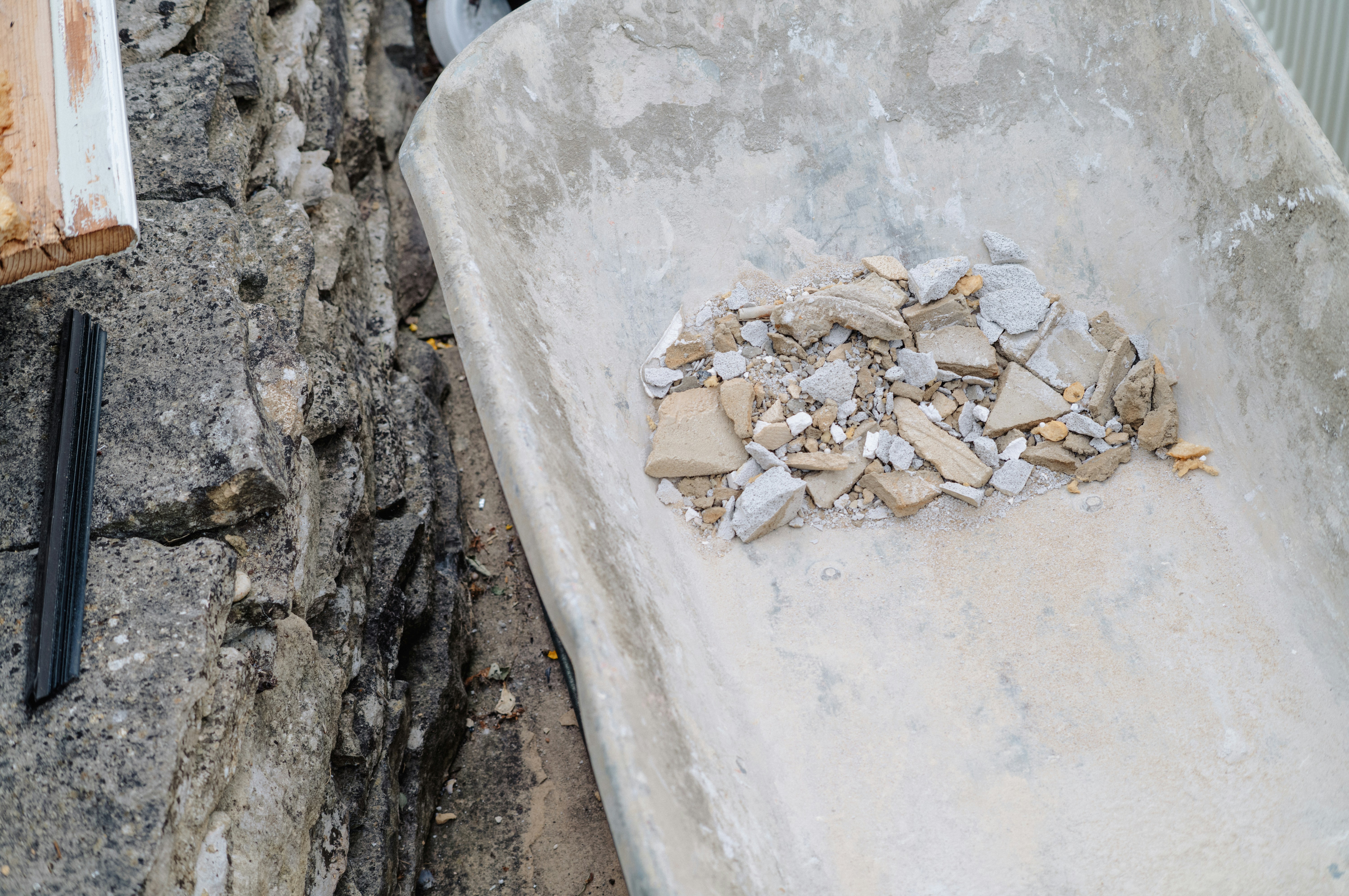 A close-up of a dusty wheelbarrow filled with broken concrete siting against a rough stone wall at a construction site.