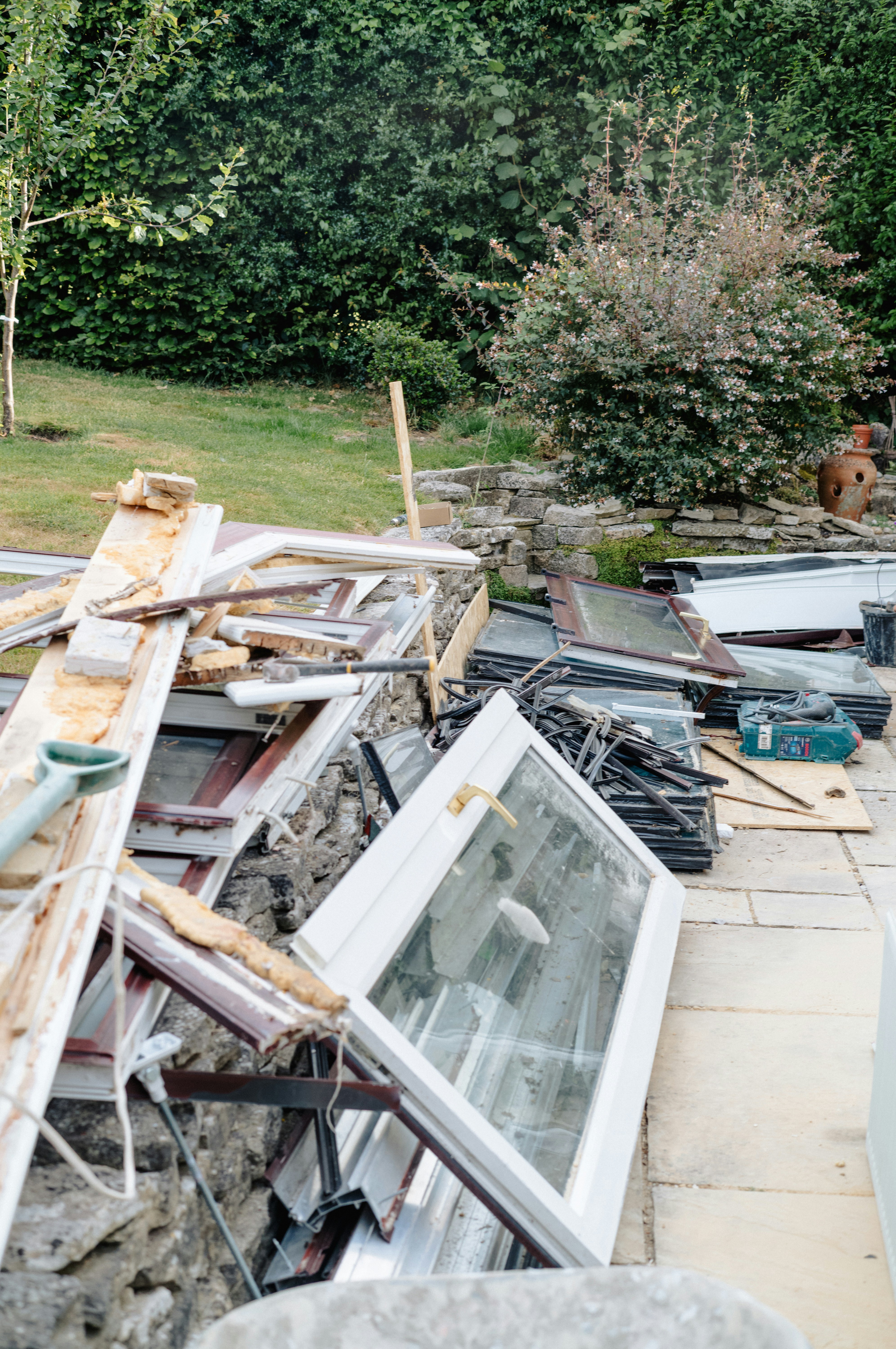 Sunlit pile of discarded window frames and glass panes alongside a stone patio, with a manicured garden and scattered debris in the background.