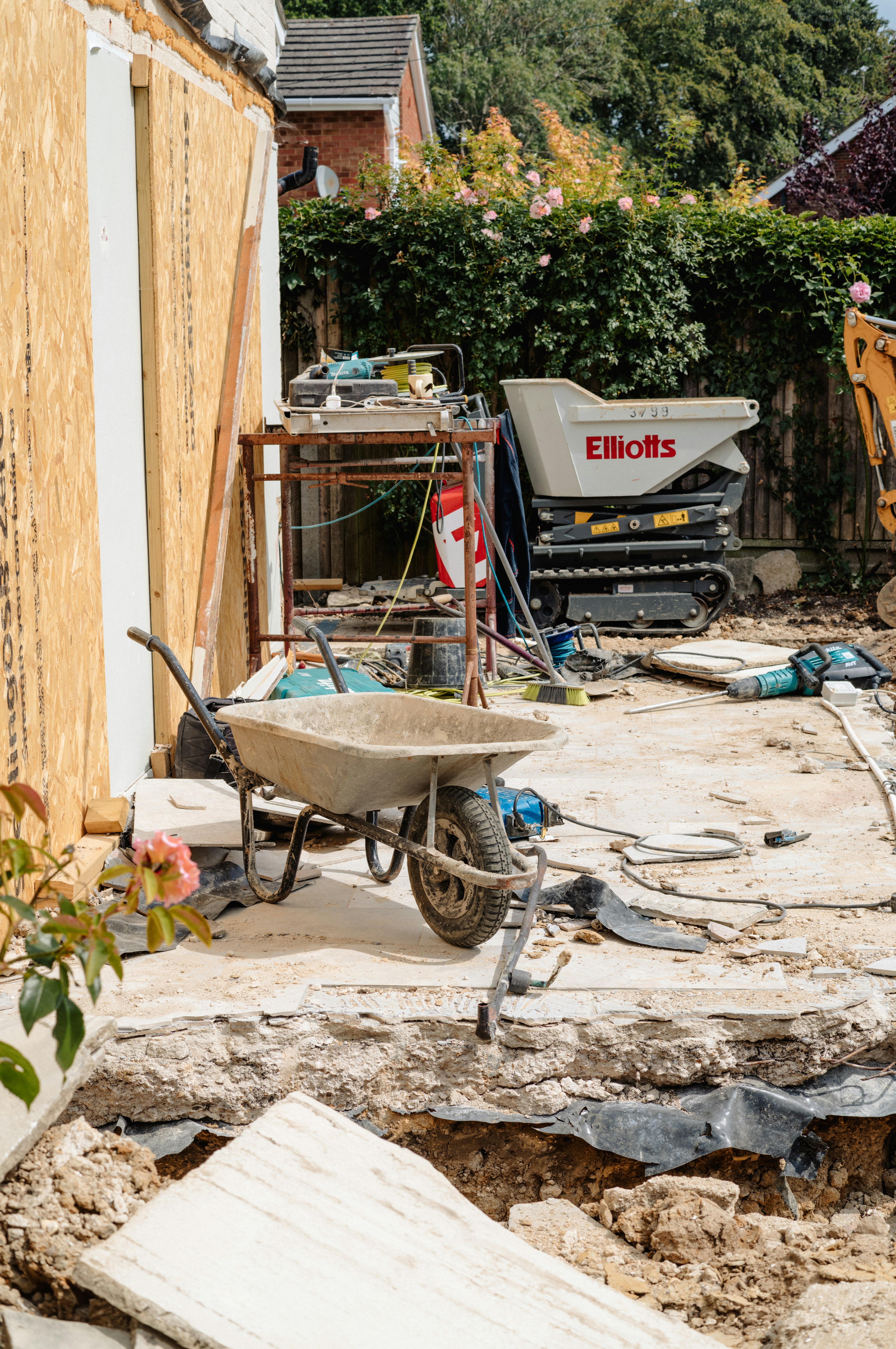 Bright, cluttered construction scene featuring a wheelbarrow in the foreground, scattered debris, and a small excavator in the background among fresh soil and scaffolding.