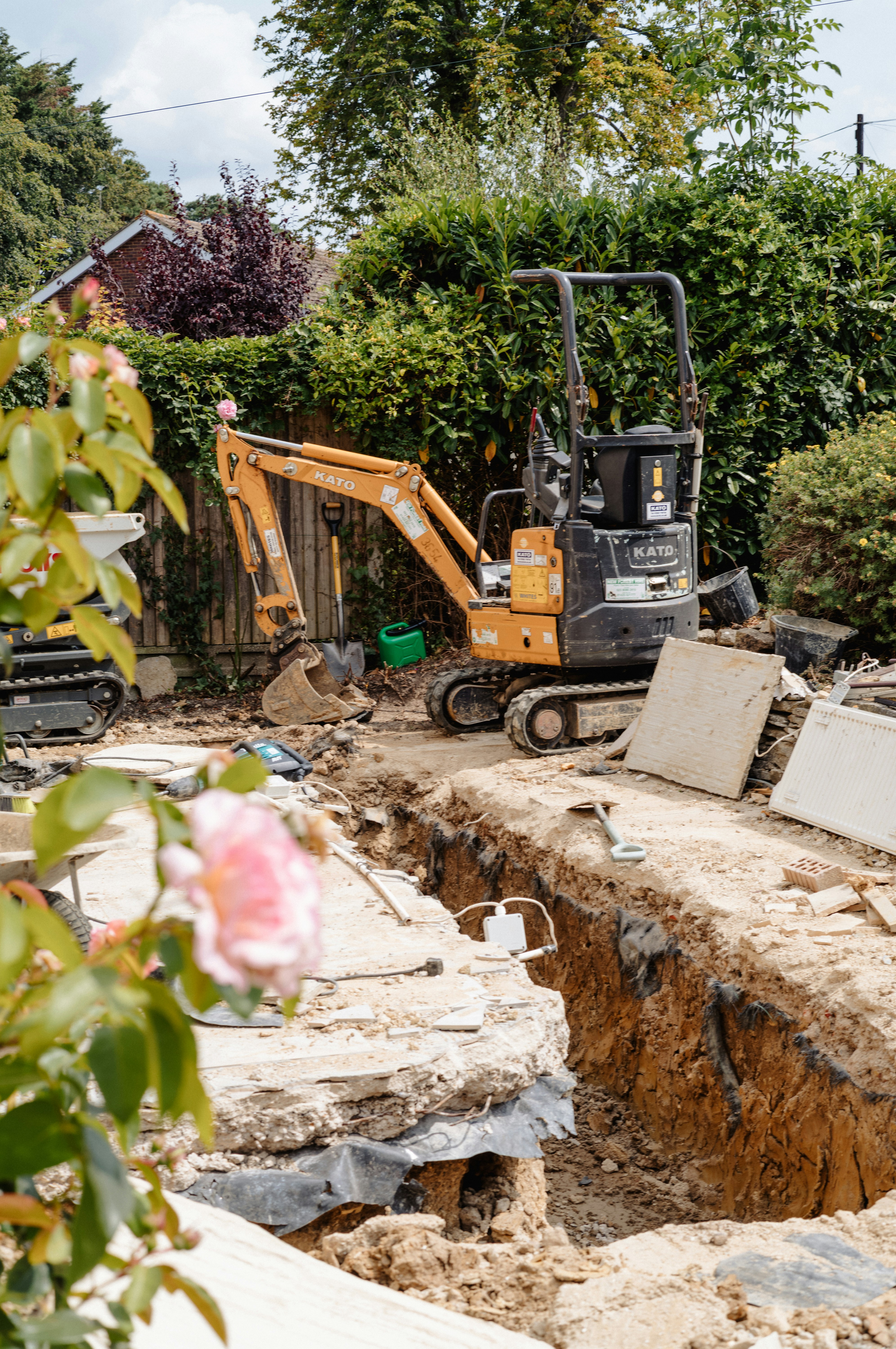 Earthmoving scene with a yellow KATO excavator at a trench site, surrounded by debris and fencing in a residential yard.