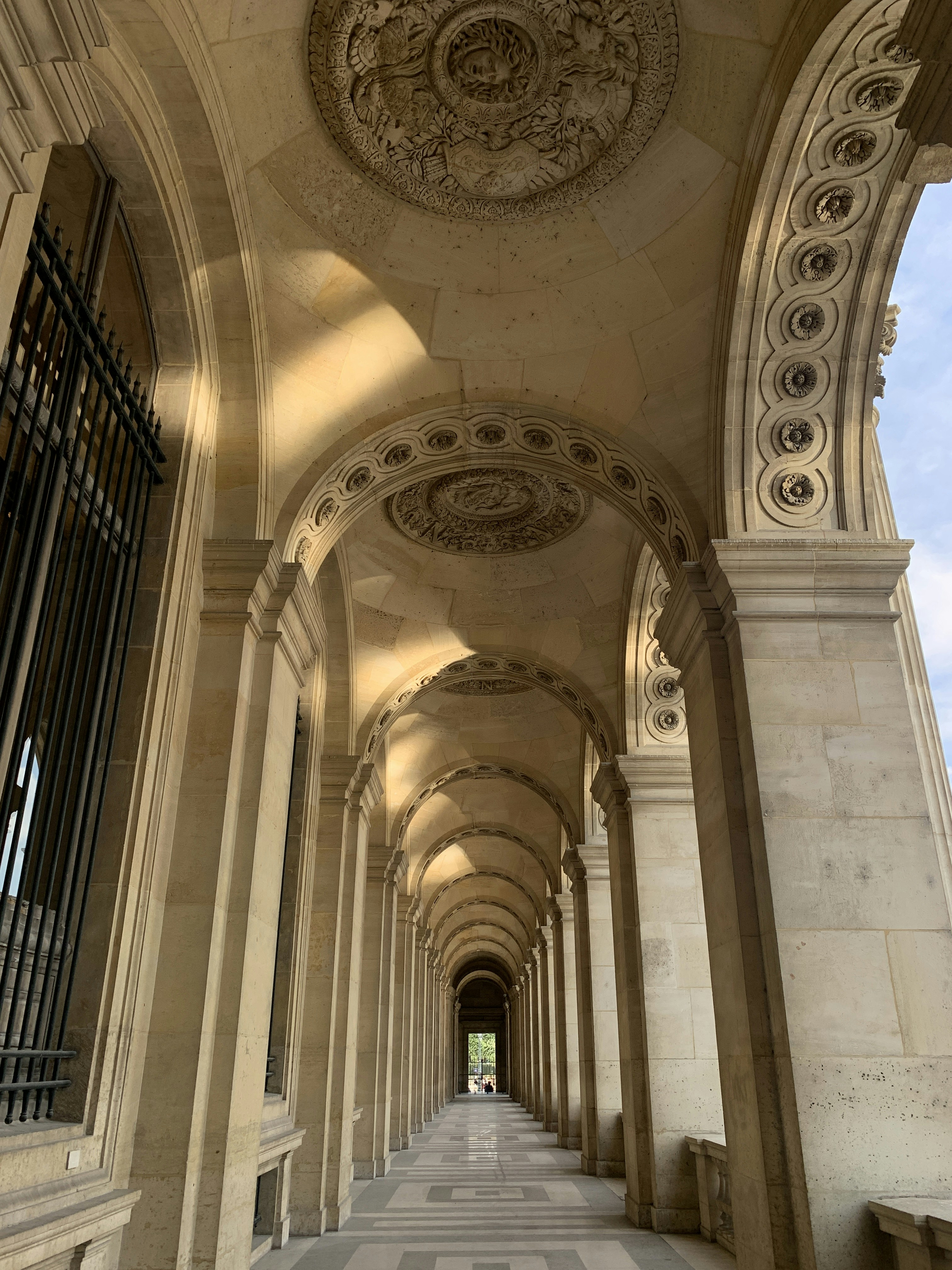 Long perspective of an arched stone colonnade with ornate ceiling medallions, leading toward a distant doorway; warm light drapes the stonework and patterned floor tiles emphasize depth.