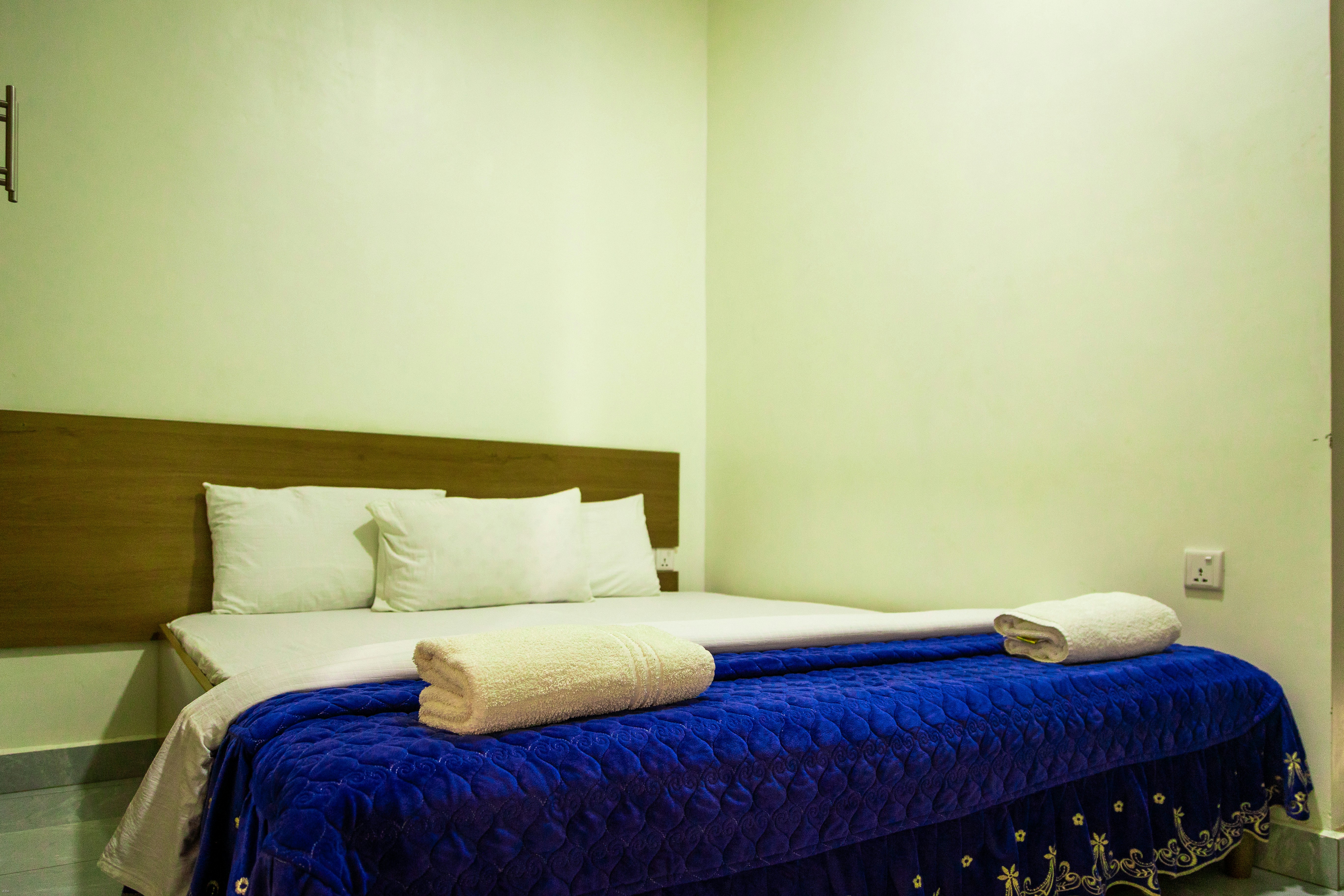 A neatly made hotel room with a double bed covered by a blue quilt and two white pillows; two rolled towels rest at the foot of the bed, set against a pale wall and simple headboard.