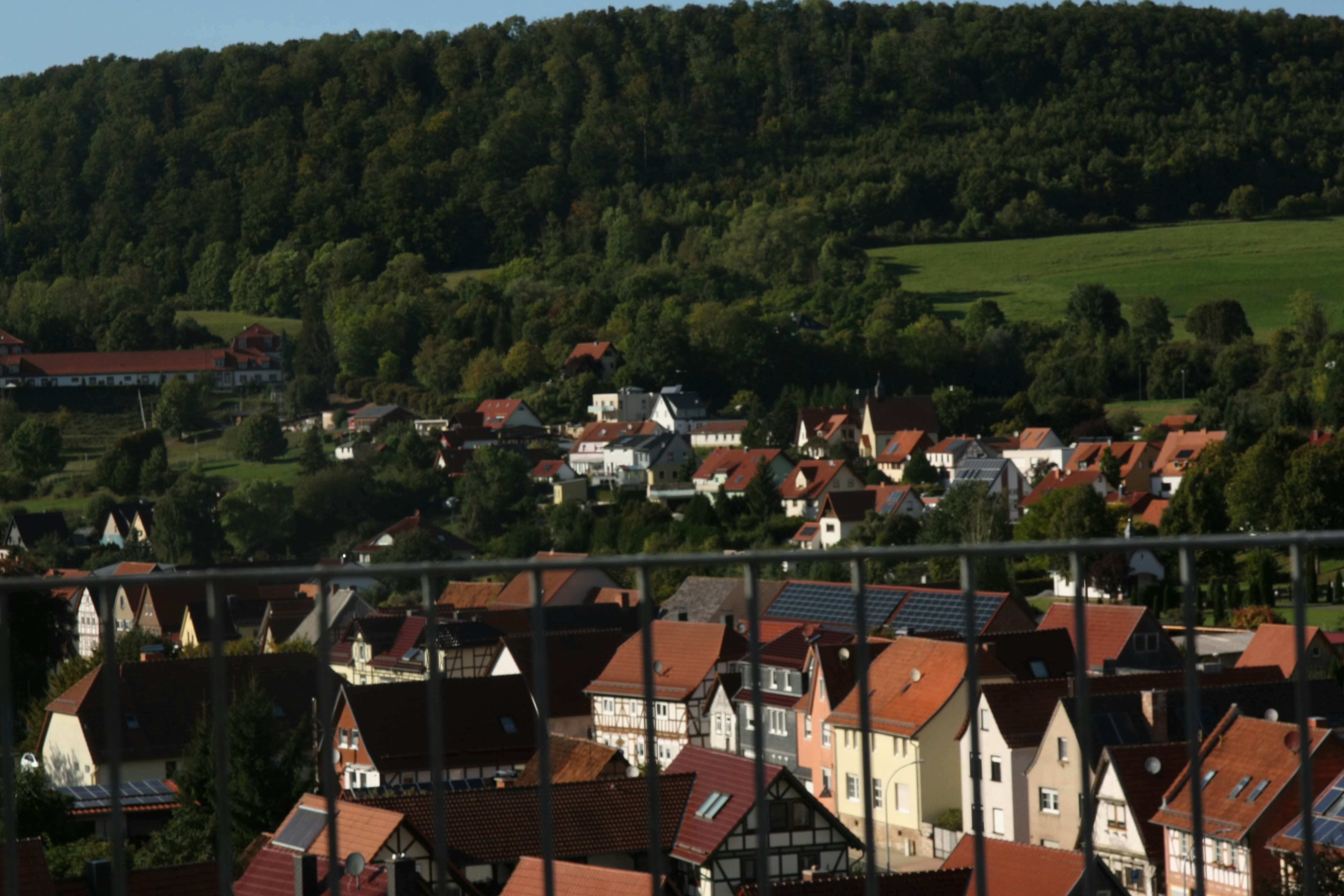 Cozy hillside town with red-tiled roofs and timber-framed facades nestled under a forested slope; a metal railing foreground hints at a balcony vantage point.