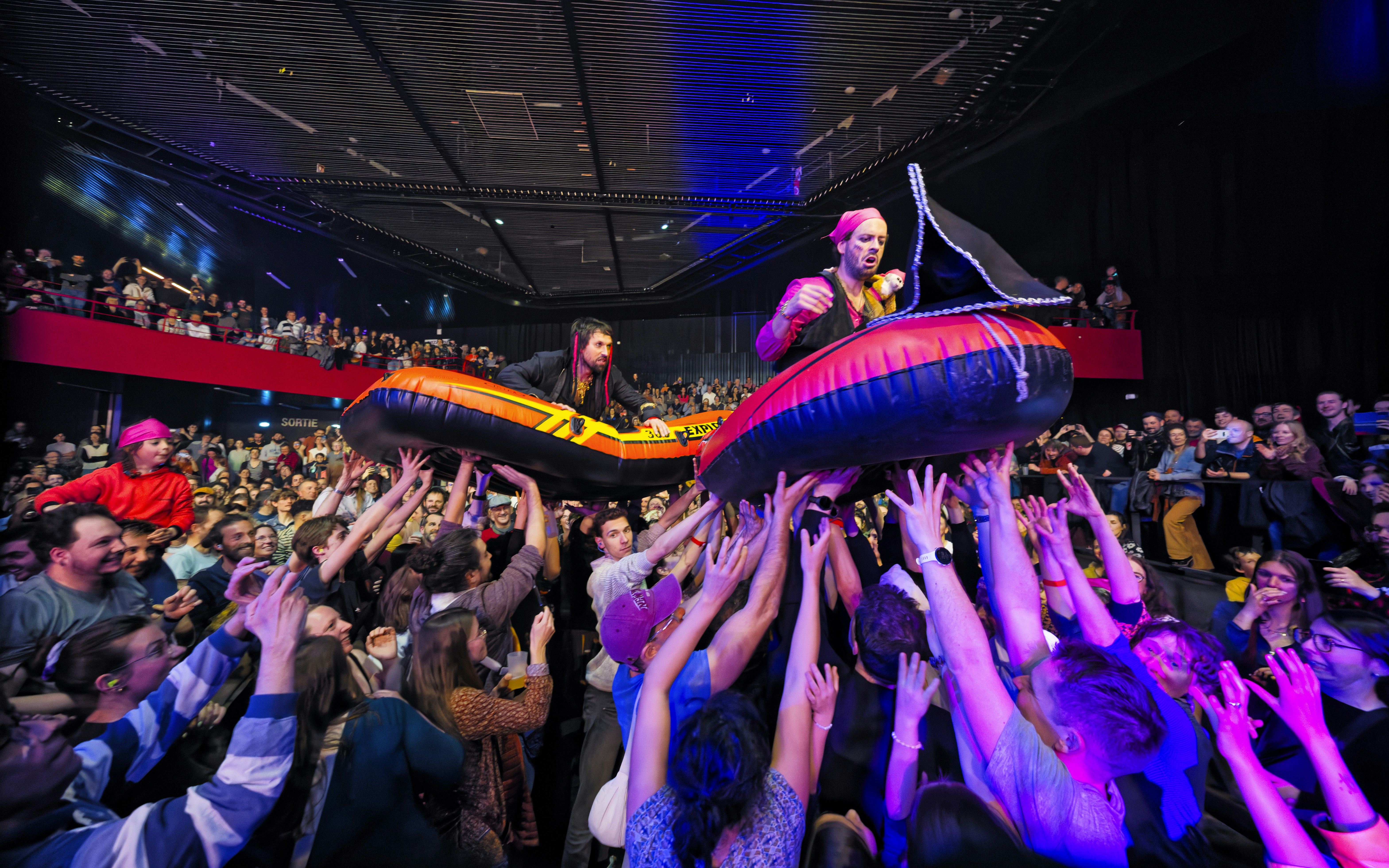 Energetic crowd surrounds performers riding an inflatable raft across the audience; venue lights bathe the scene in color while spectators reach upward.