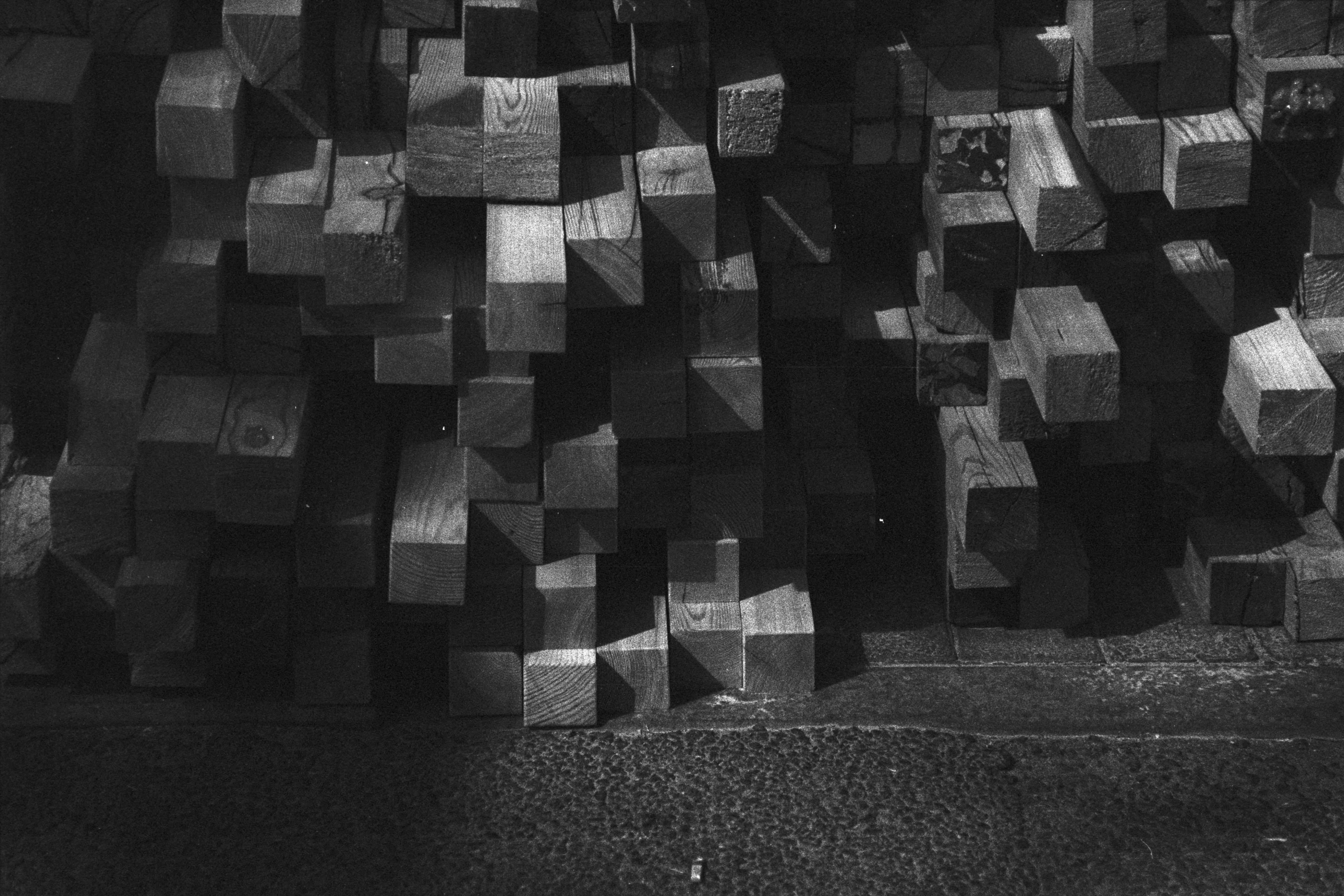 Monochrome stack of rectangular timbers arranged in a dense wall, with a rough ground plane at the base. The composition emphasizes texture and geometric repetition over color or human presence.