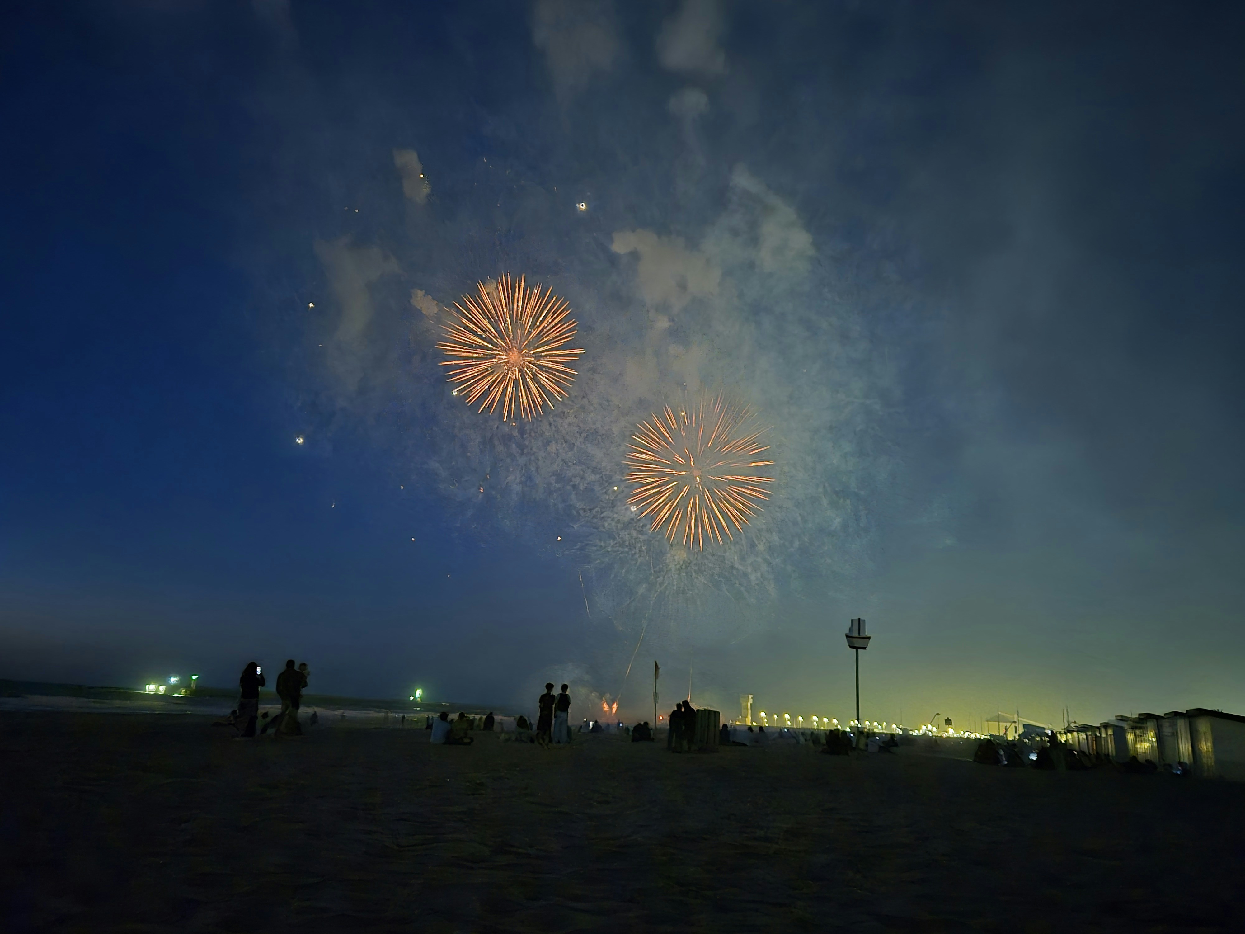 Two large fireworks blooms illuminate the night sky above a dark beach, with small silhouettes of people and distant city lights along the horizon.