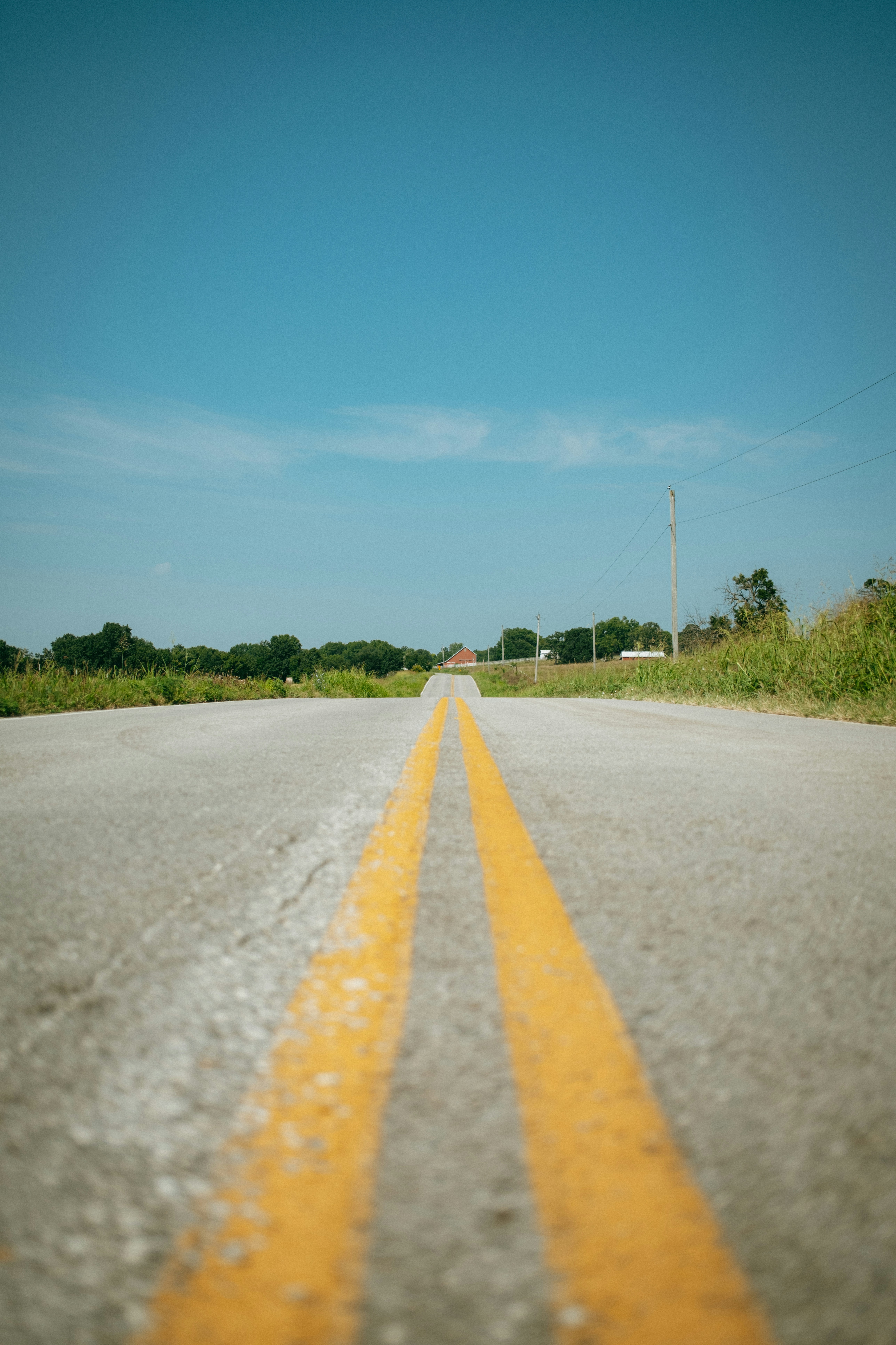 Asphalt road with double yellow lines stretches toward a distant vanishing point under a clear blue sky; rural landscape on either side, power lines running along the road.