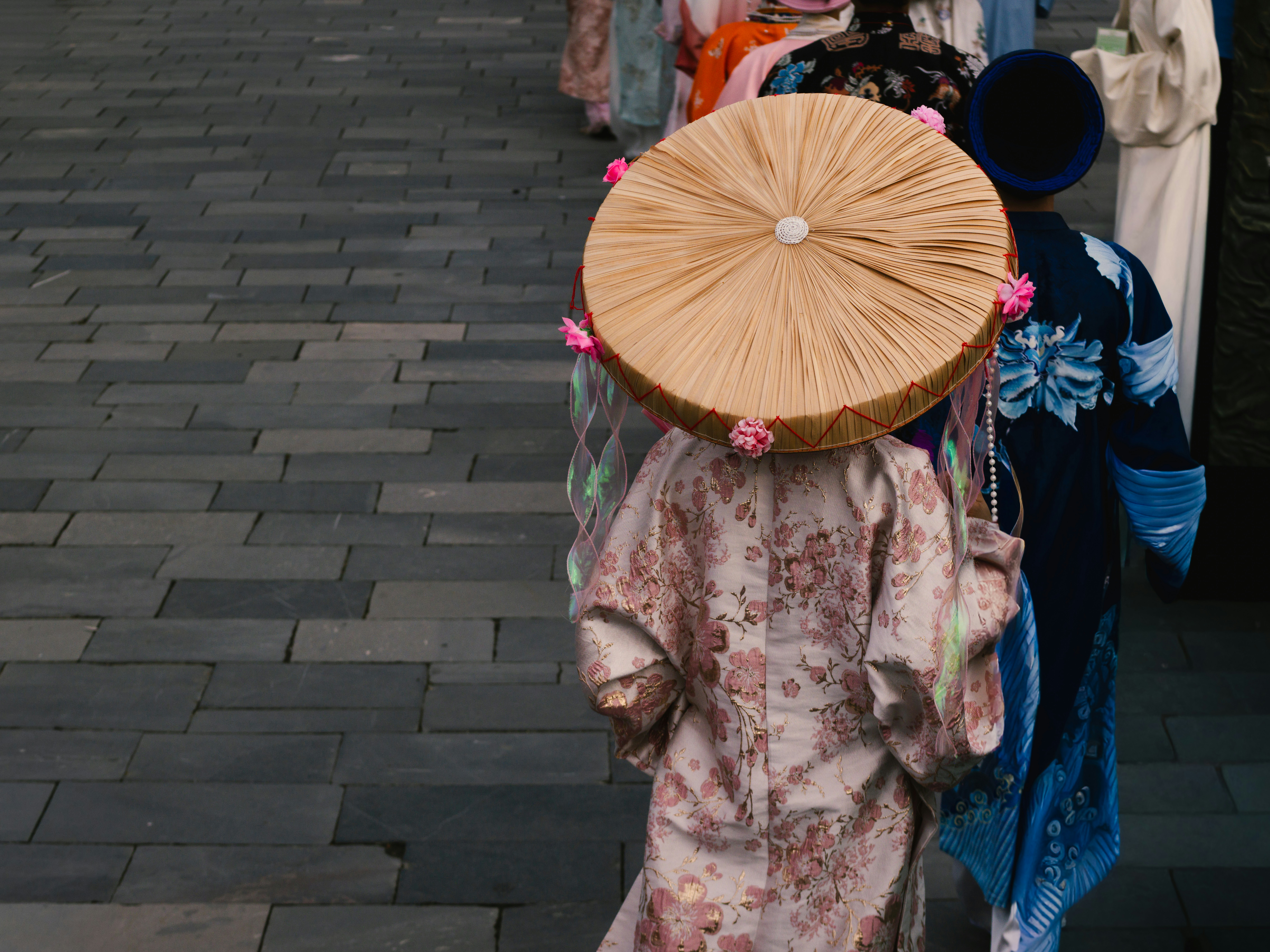 Colorful street scene featuring a person wearing a traditional kimono-style robe and a wide, straw hat, viewed from behind as they walk among others on a paved walkway.