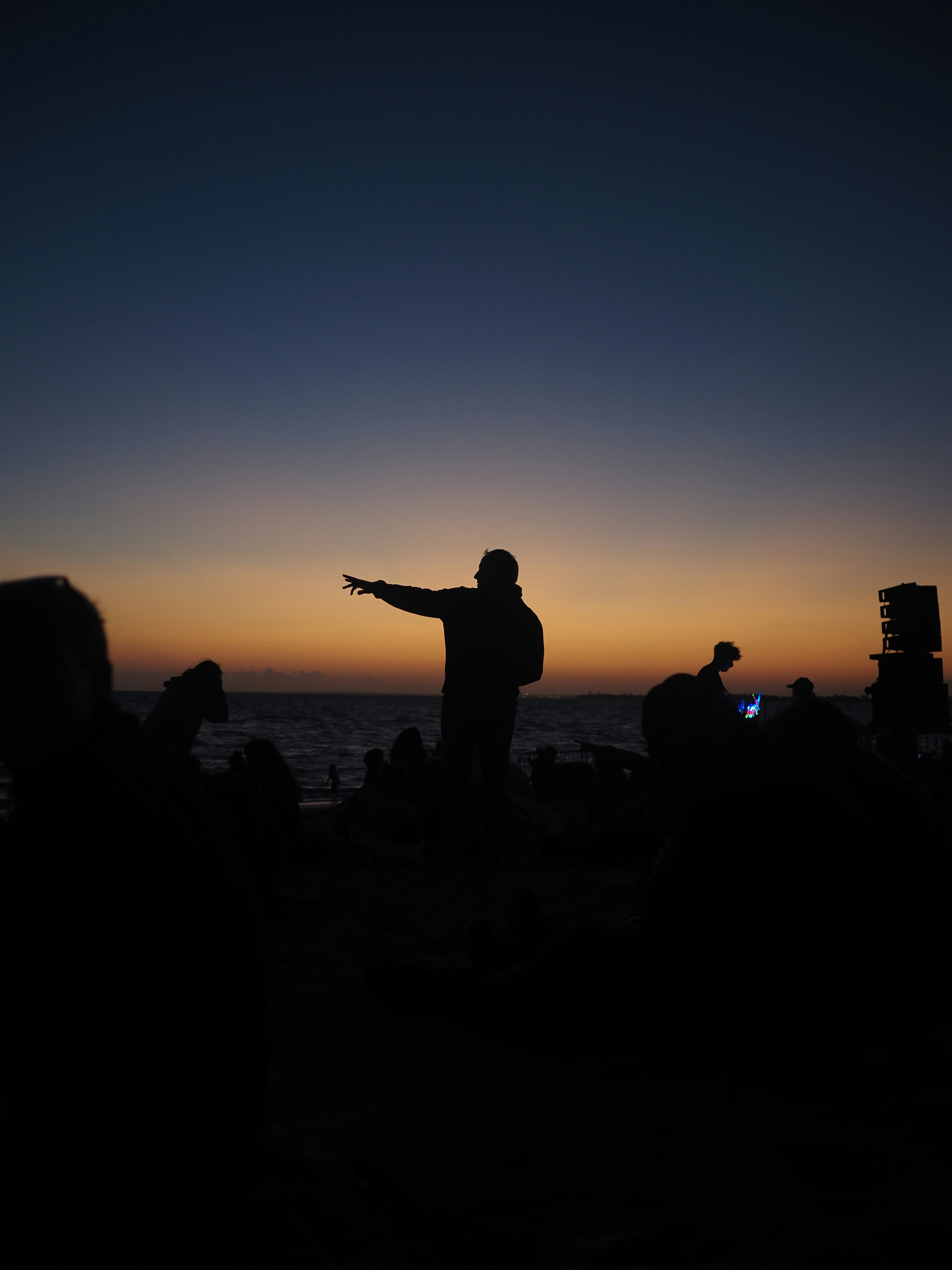 Silhouetted figures gather along a beach at sunset, a central speaker extends an arm toward the sea while distant light accents hint at a performance setup.