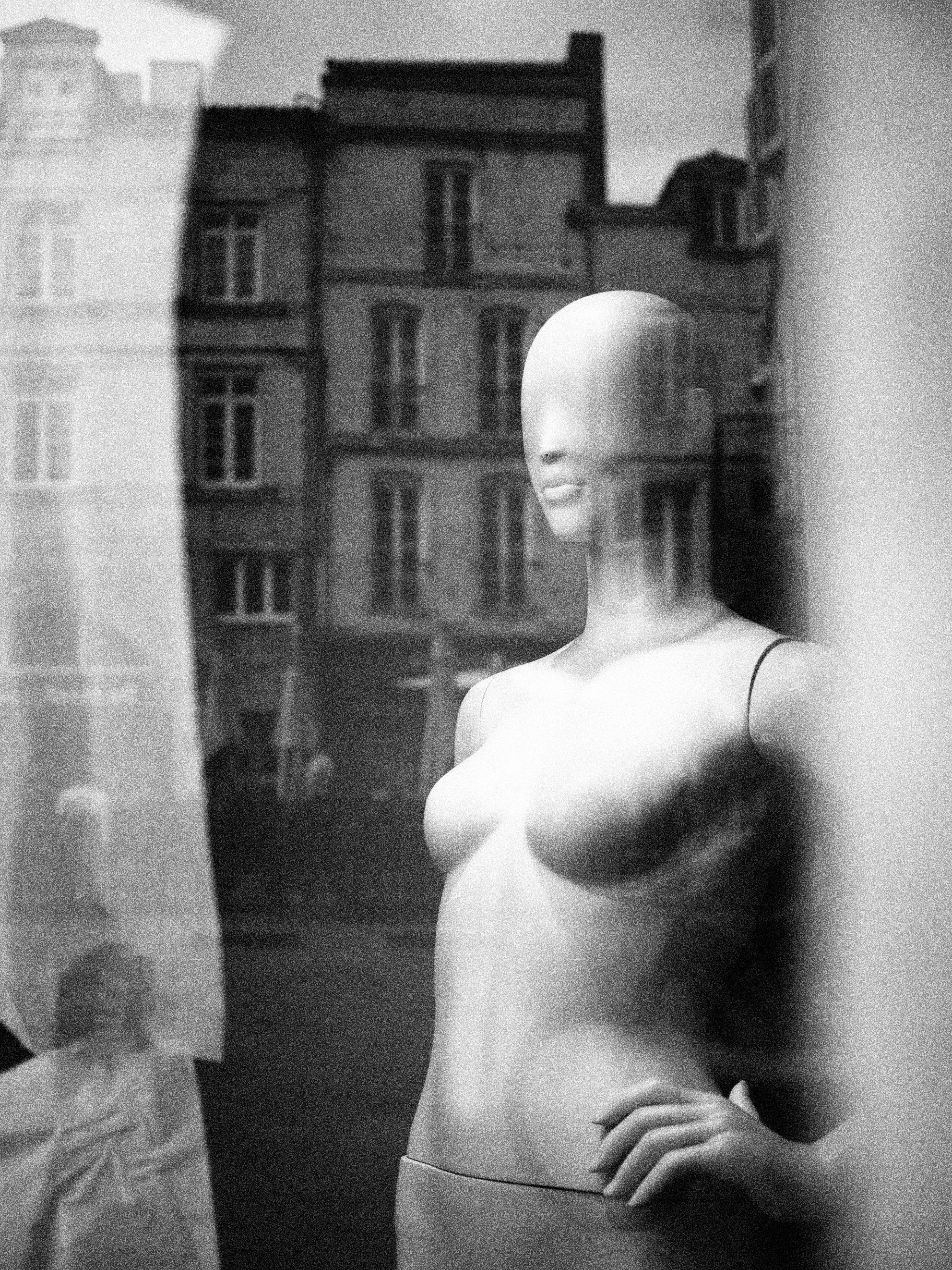 Monochrome mannequin torso in a shop window with reflected urban buildings, creating a double-exposure effect and a silent, architectural portrait.
