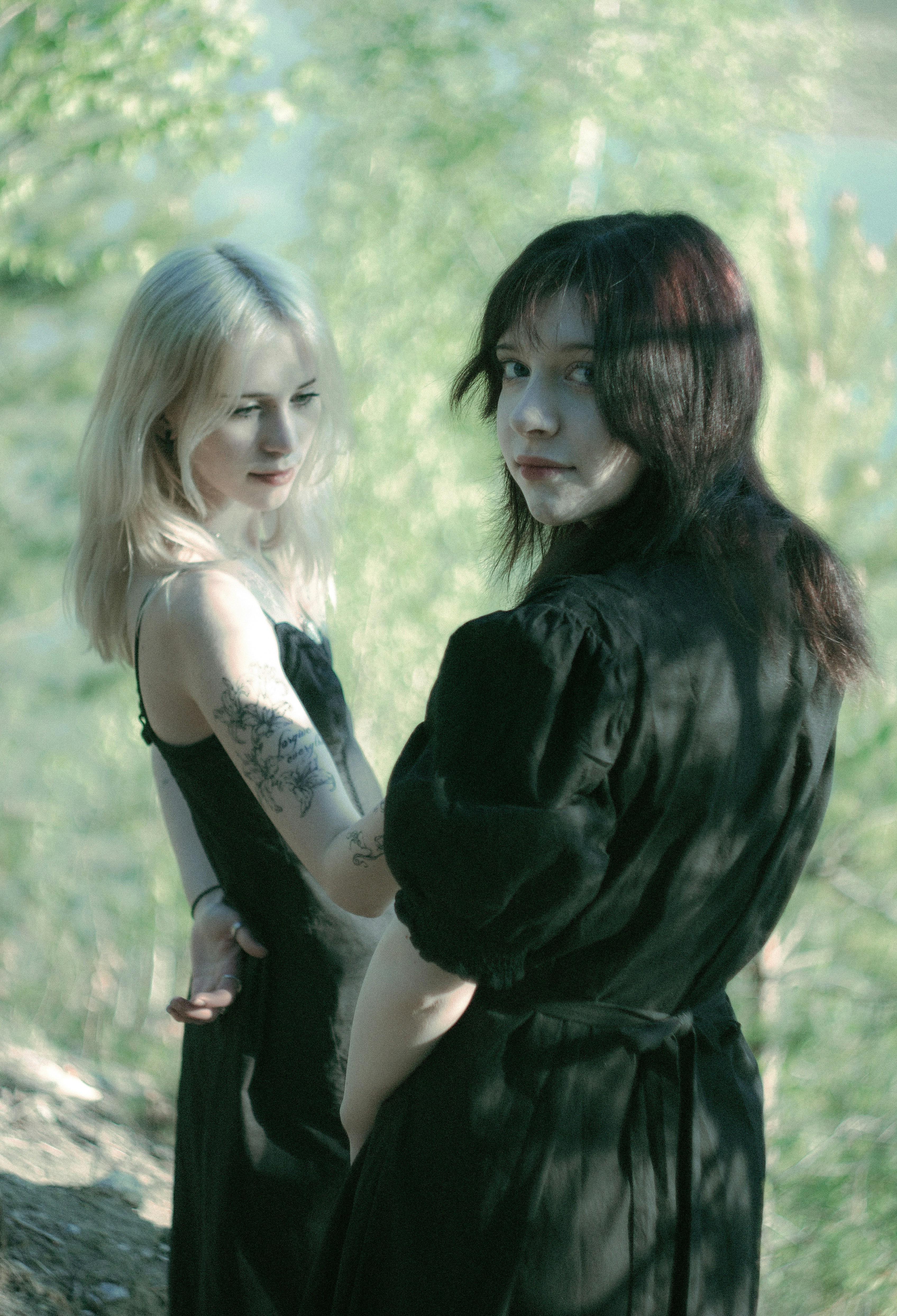 Two friends in a sunlit forest, dressed in black, sharing a moment of quiet connection