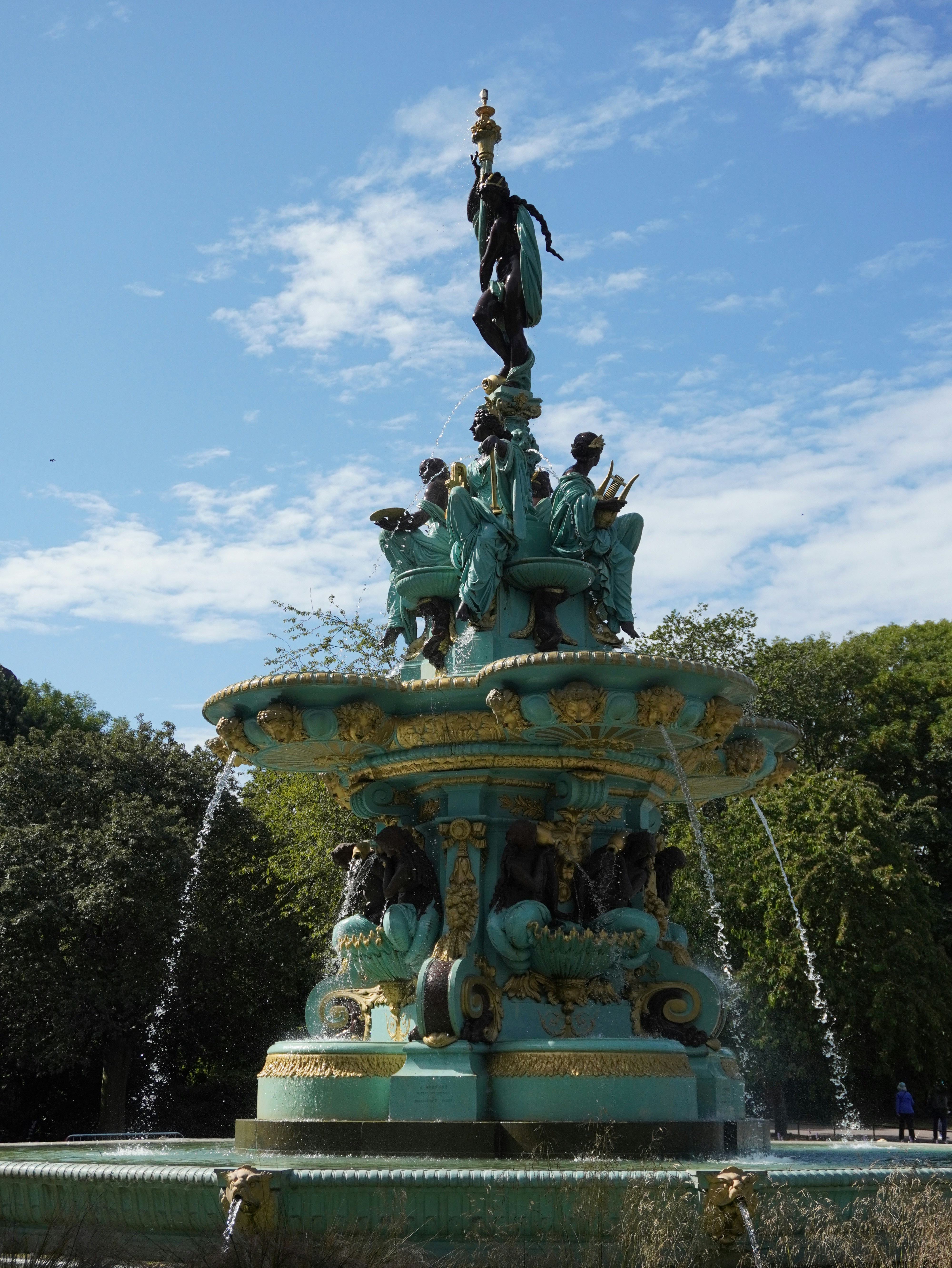 A turquoise multi-tier fountain sculpture rises against a bright blue sky with trees in the background, capturing ornate details and cascading water. This is a water-fountain monument.