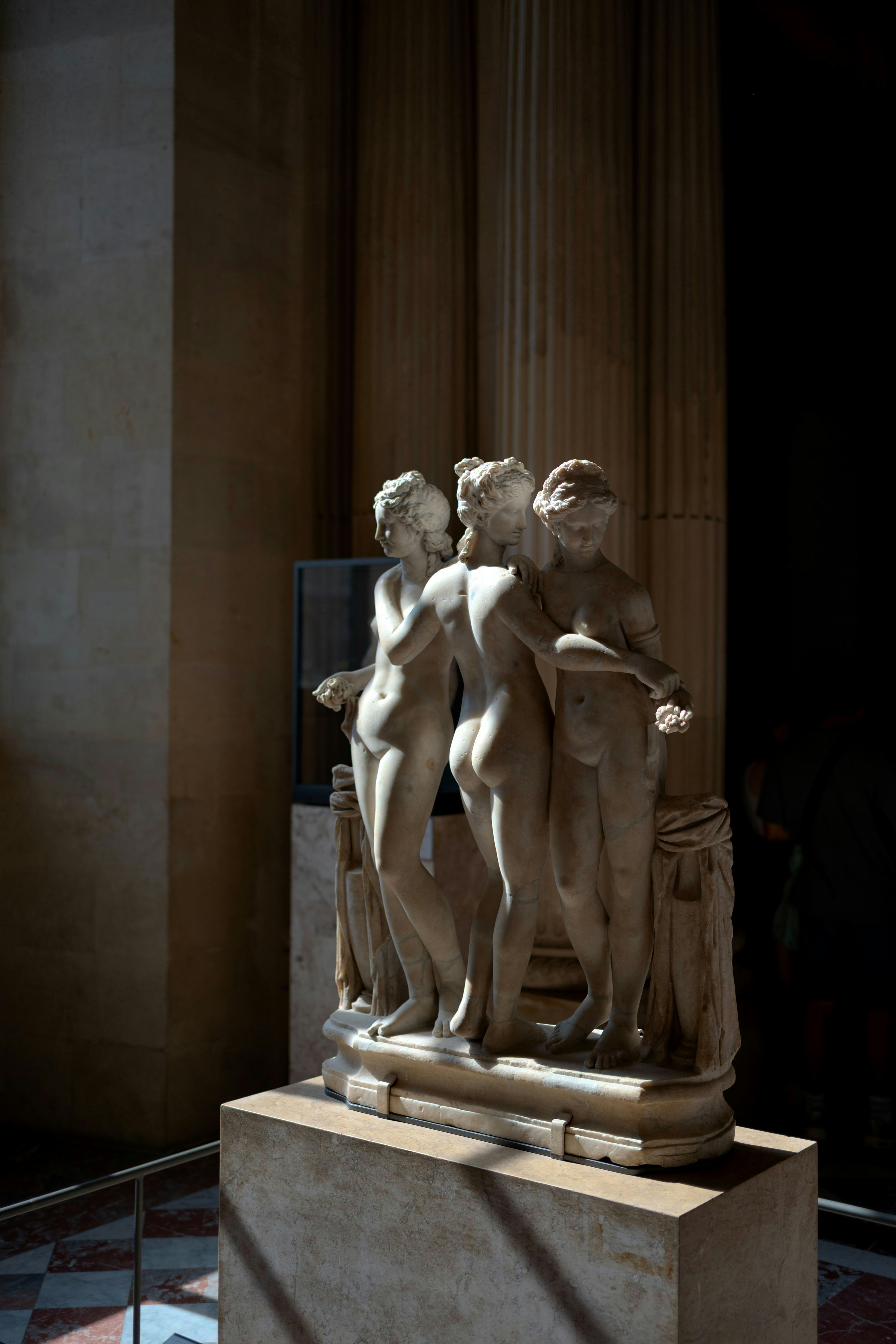 White marble sculpture of three intertwined figures on a pedestal in a dim museum hall; soft spotlight emphasizes their contours and creates deep shadows.