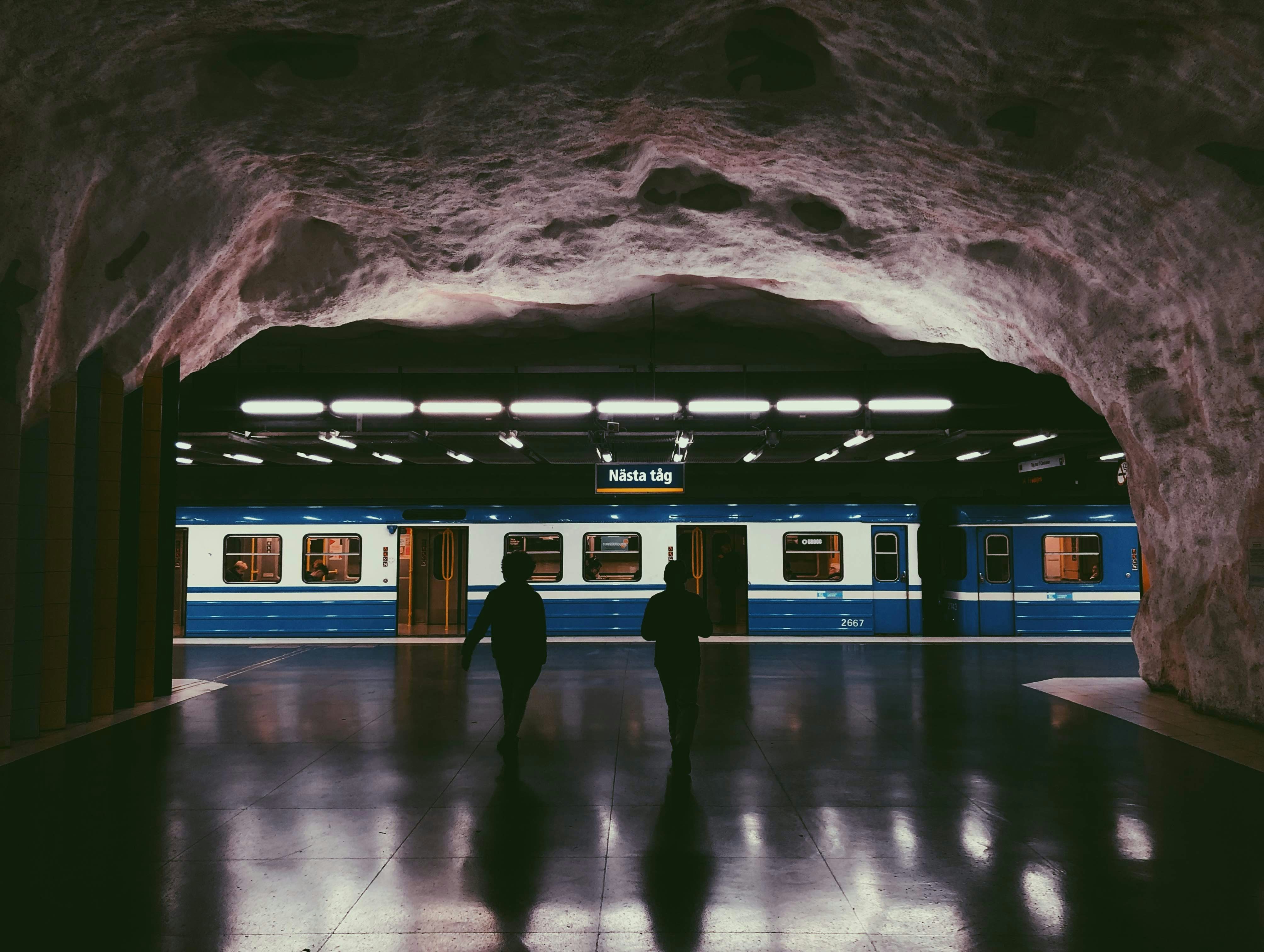 Two silhouetted figures stand in a dim underground station beneath a curved, rocky arch, with a blue train at the platform and reflections on the polished floor.
