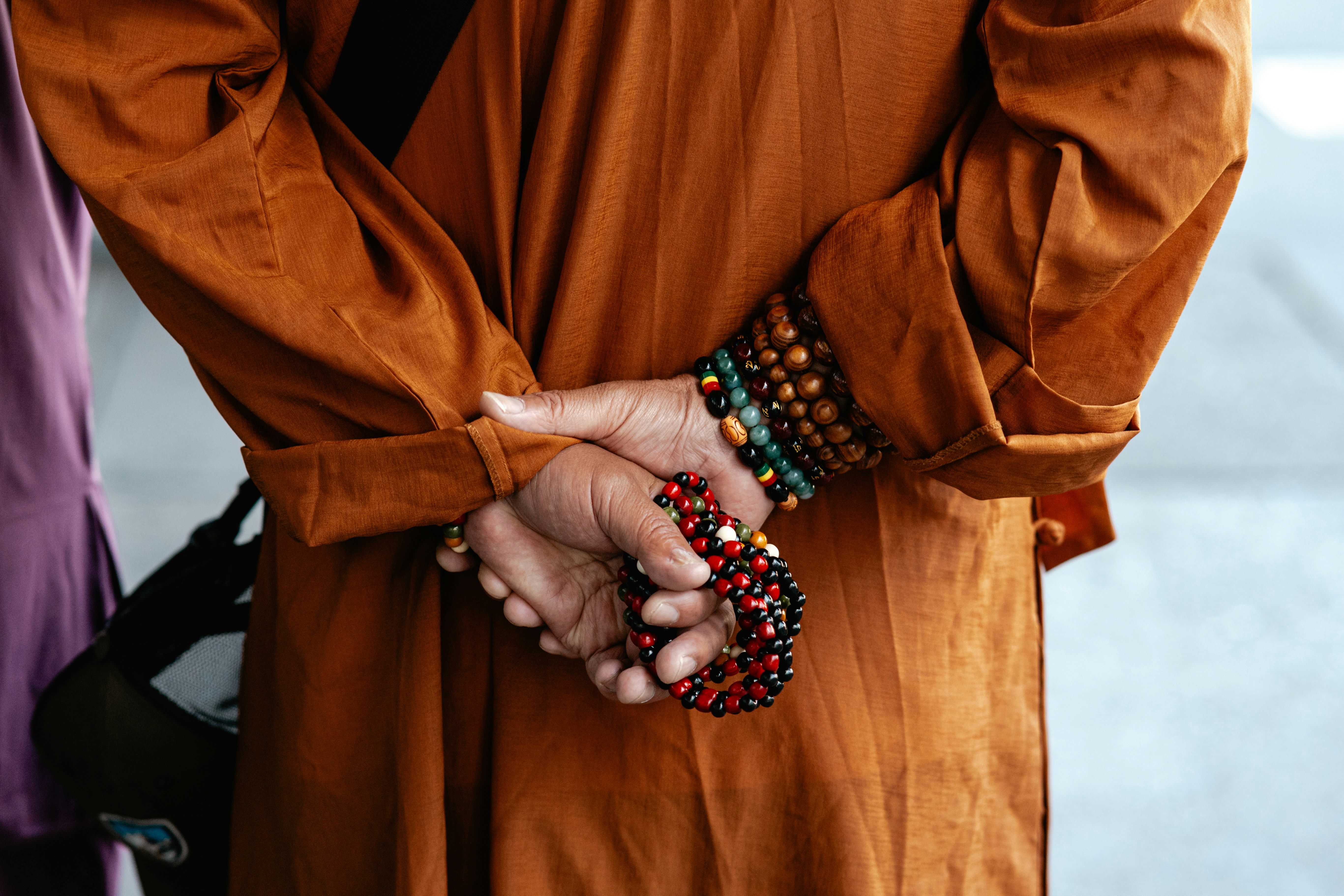 Close-up of hands clasped behind an amber robe, adorned with multicolored bead bracelets; a glimpse of purple fabric and a shoulder strap in the background.