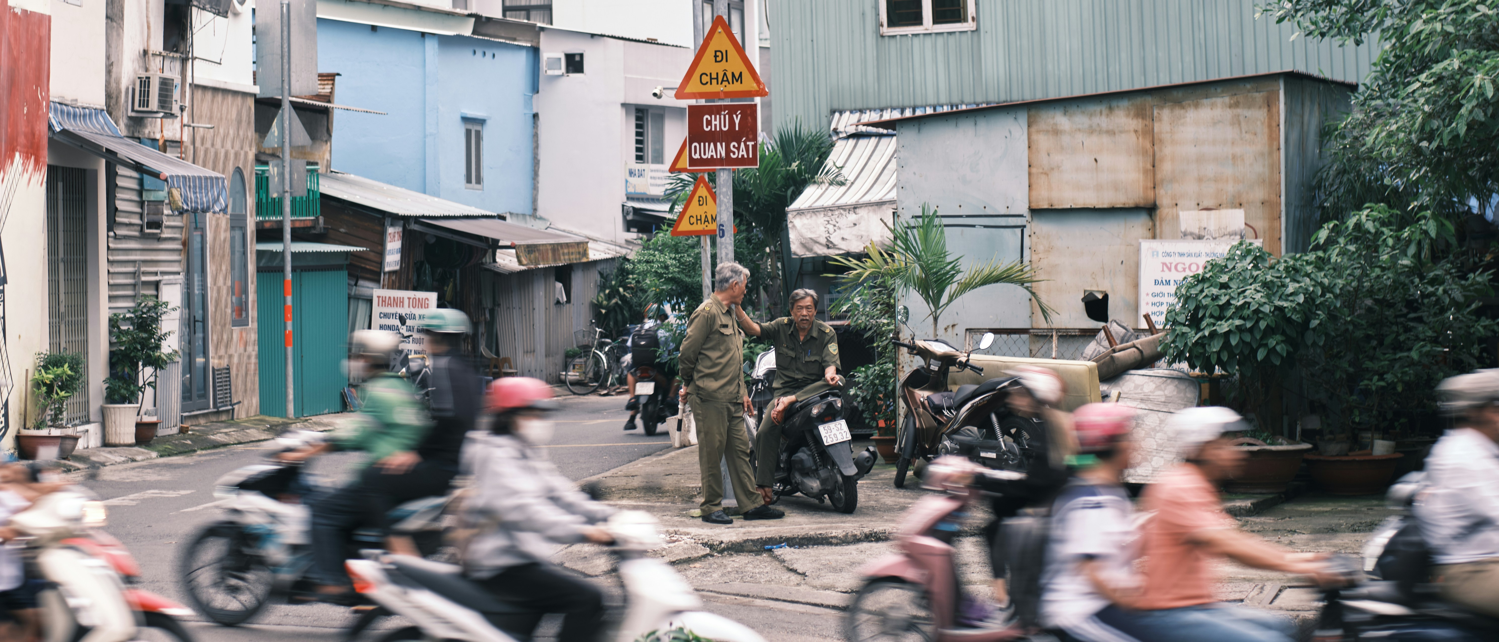 Bustling street scene with motorbikes blurred in motion and a line of guards or officials near a pedestrian crossing; urban storefronts and urban signage in the background, conveying everyday city life.
