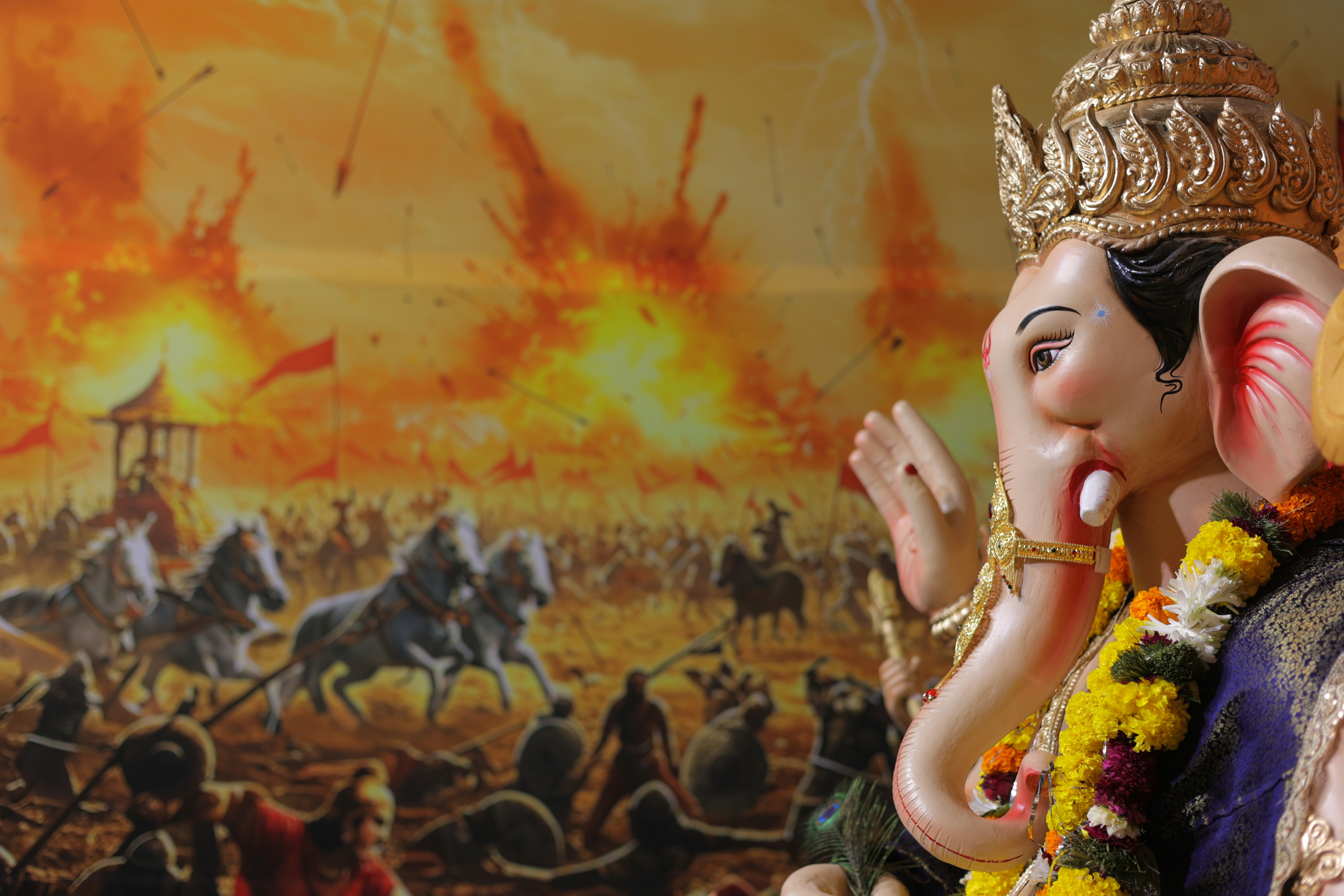 Close-up of a decorated Ganesha statue with garlands, set against a dramatic fiery battle mural in the background.