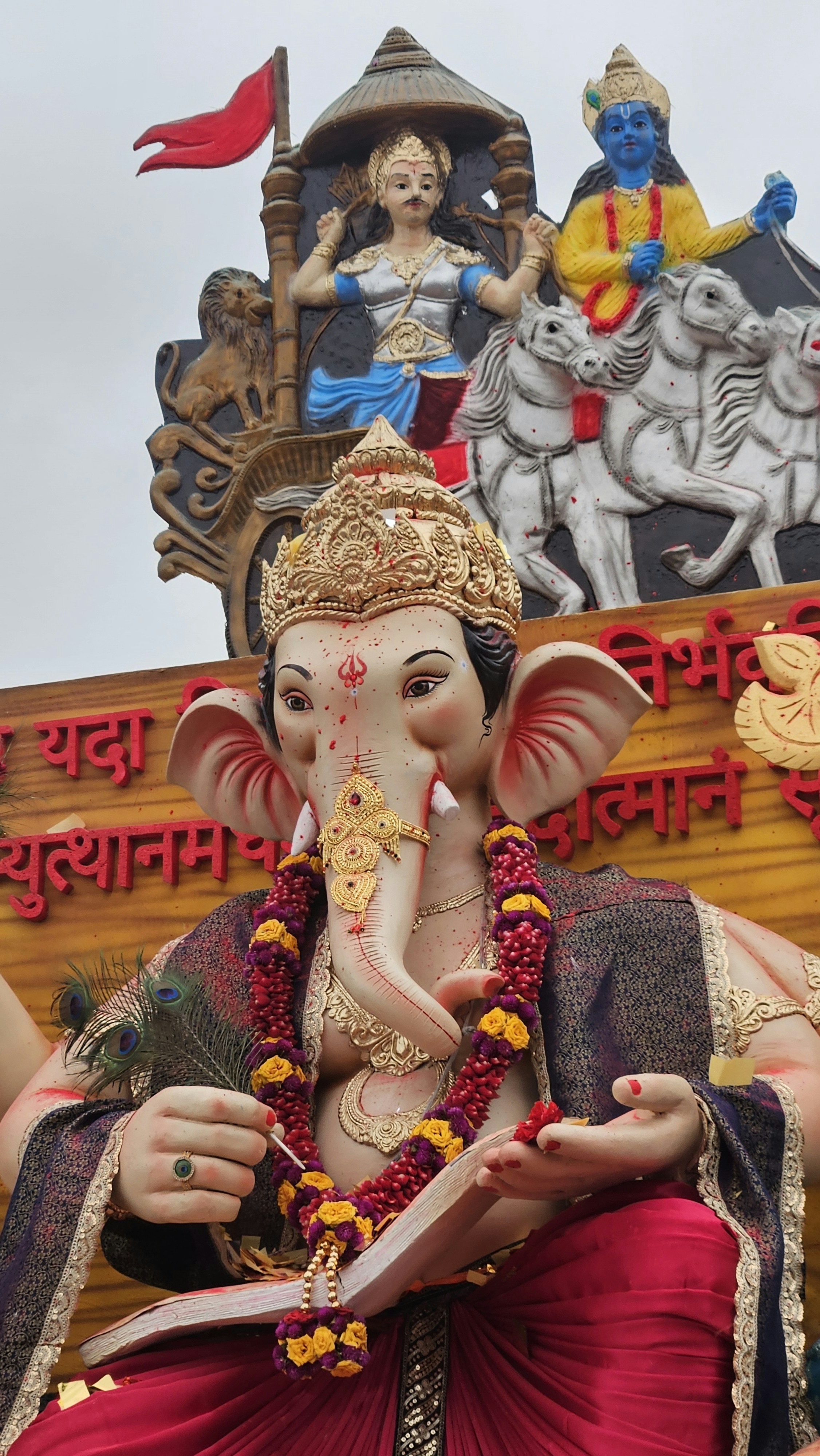 Vibrant Ganesha statue with a bejeweled crown and garlands, flanked by a decorative backdrop of mythic riders on horses during a festival scene.