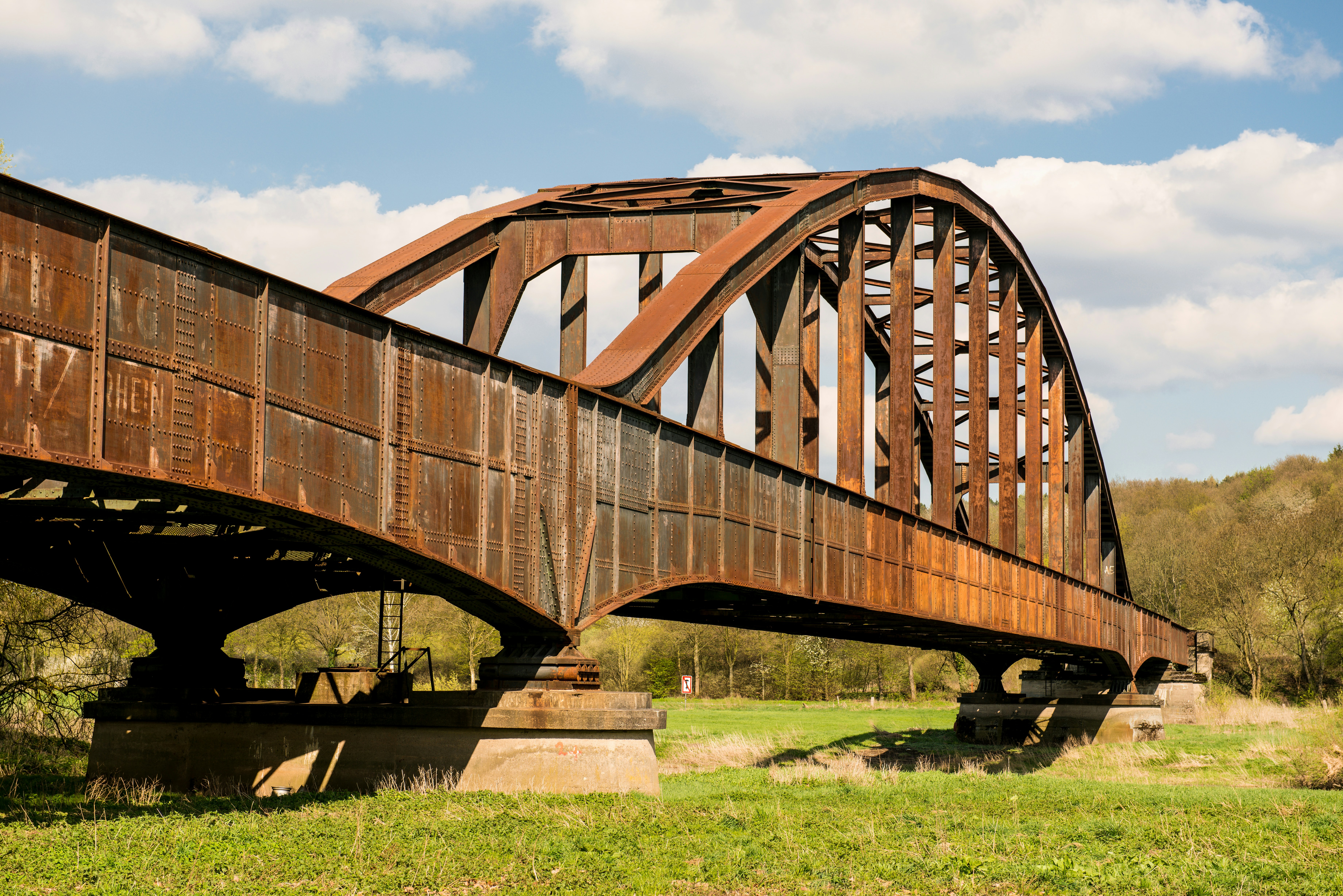 Rusty iron arch bridge spanning a grassy riverbed with a clear sky and scattered trees in the background.
