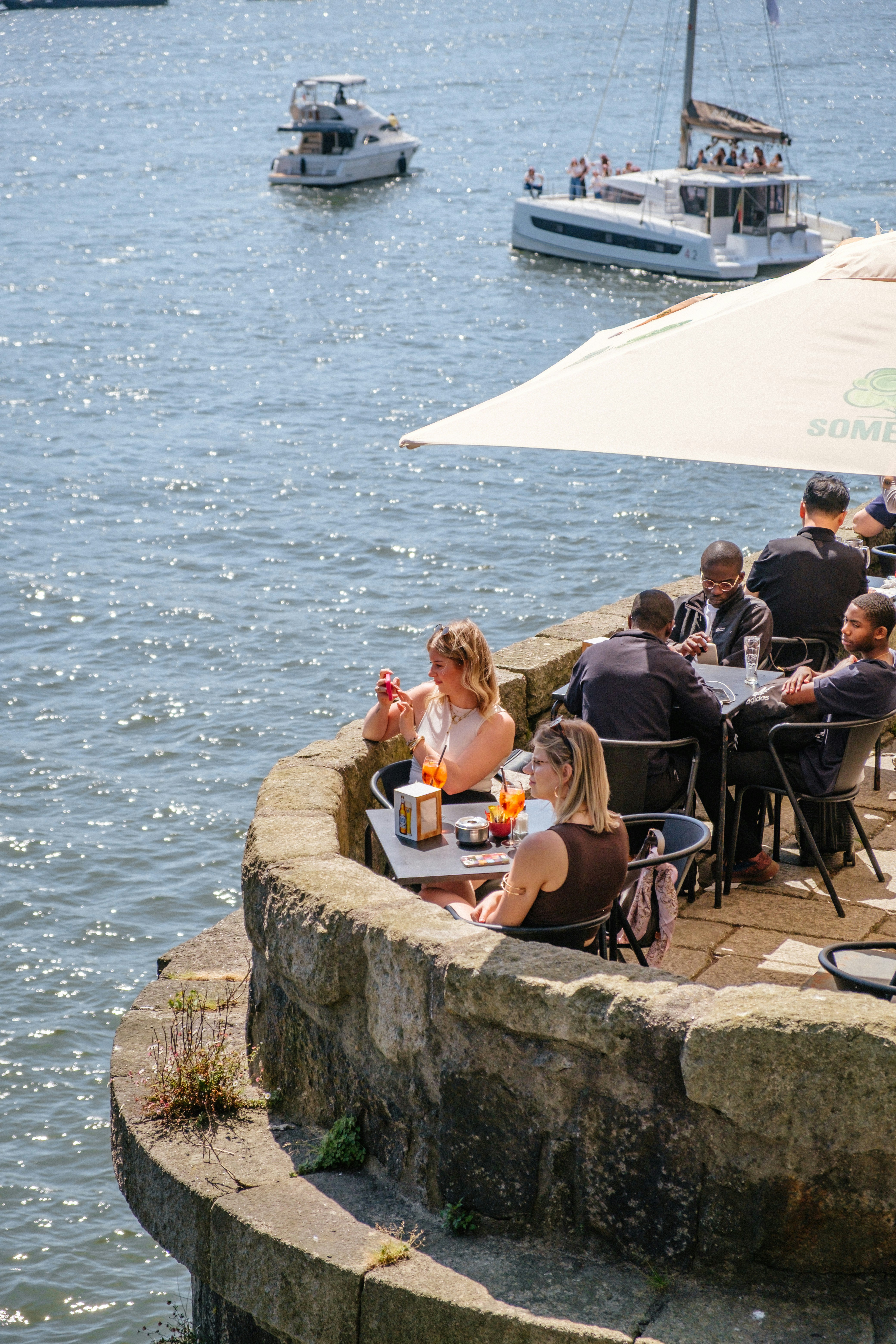 Diners sit at an outdoor stone terrace by the water, sipping drinks as boats drift on a sunlit harbor behind them. A large umbrella shades part of the scene, emphasizing the seaside dining vibe.
