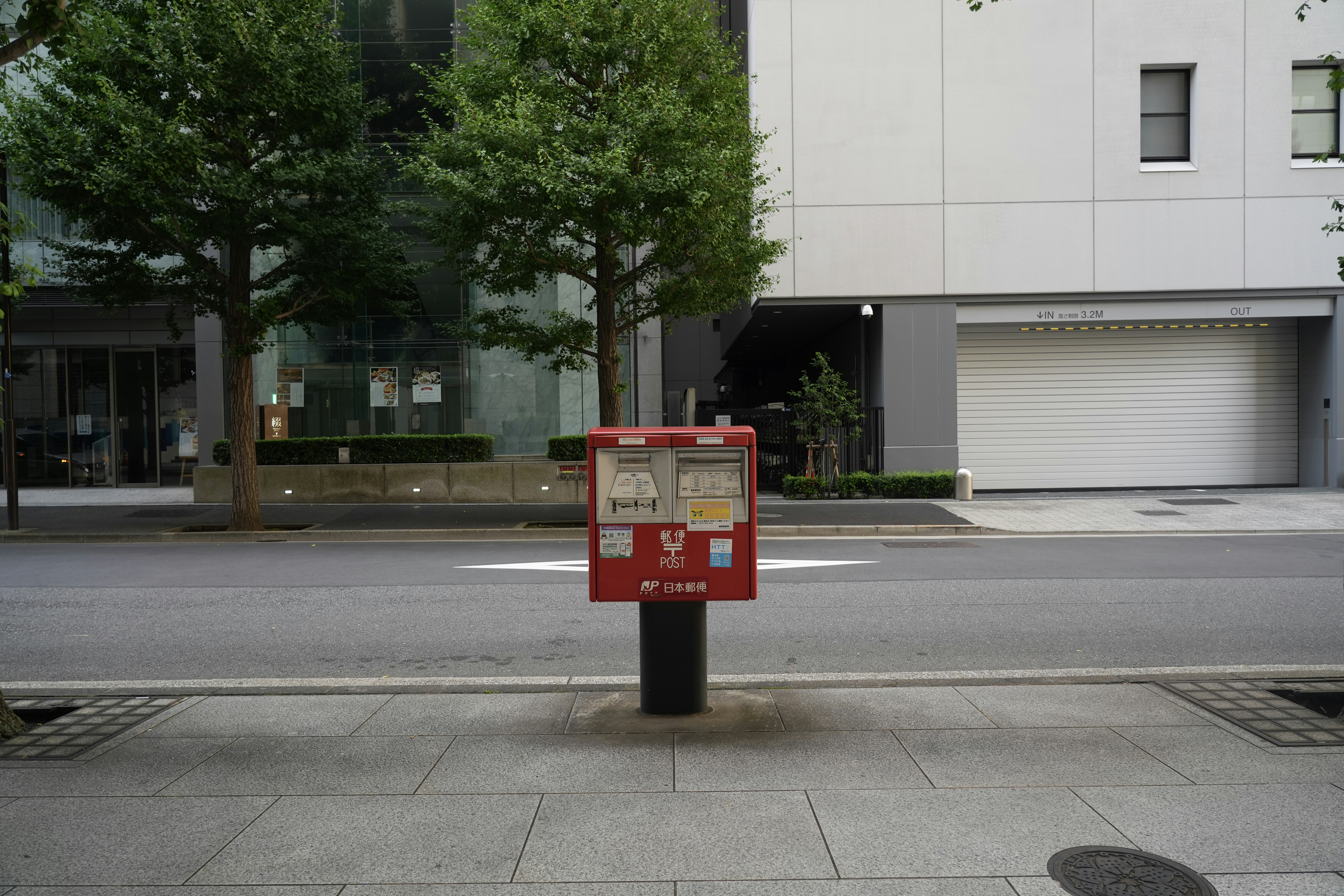 A solitary red mailbox stands on a paved urban sidewalk with modern buildings and trees in the background, conveying everyday city life.