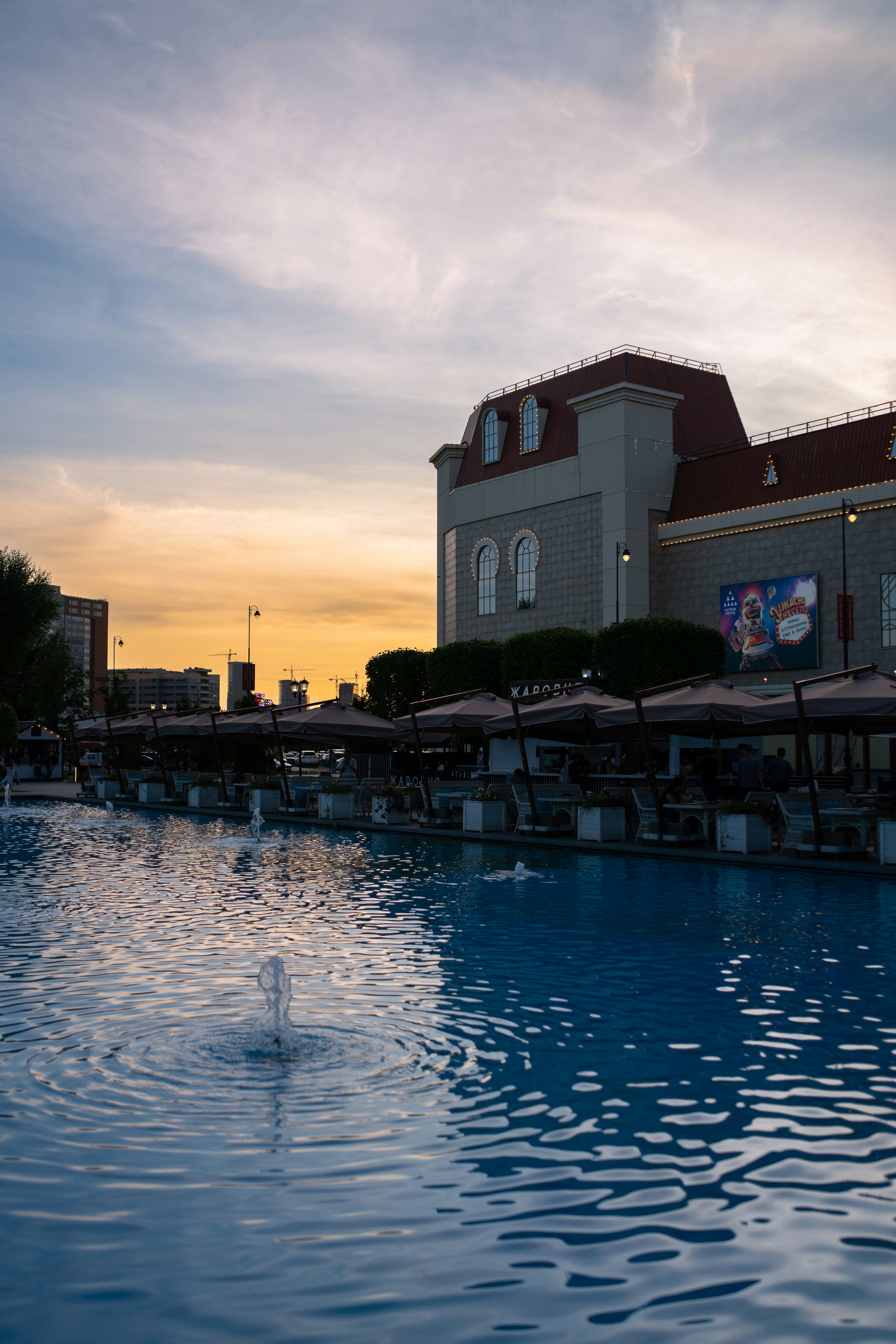 Golden sunset casts warm light over a hotel pool courtyard with umbrellas and a grand building facade in the background.