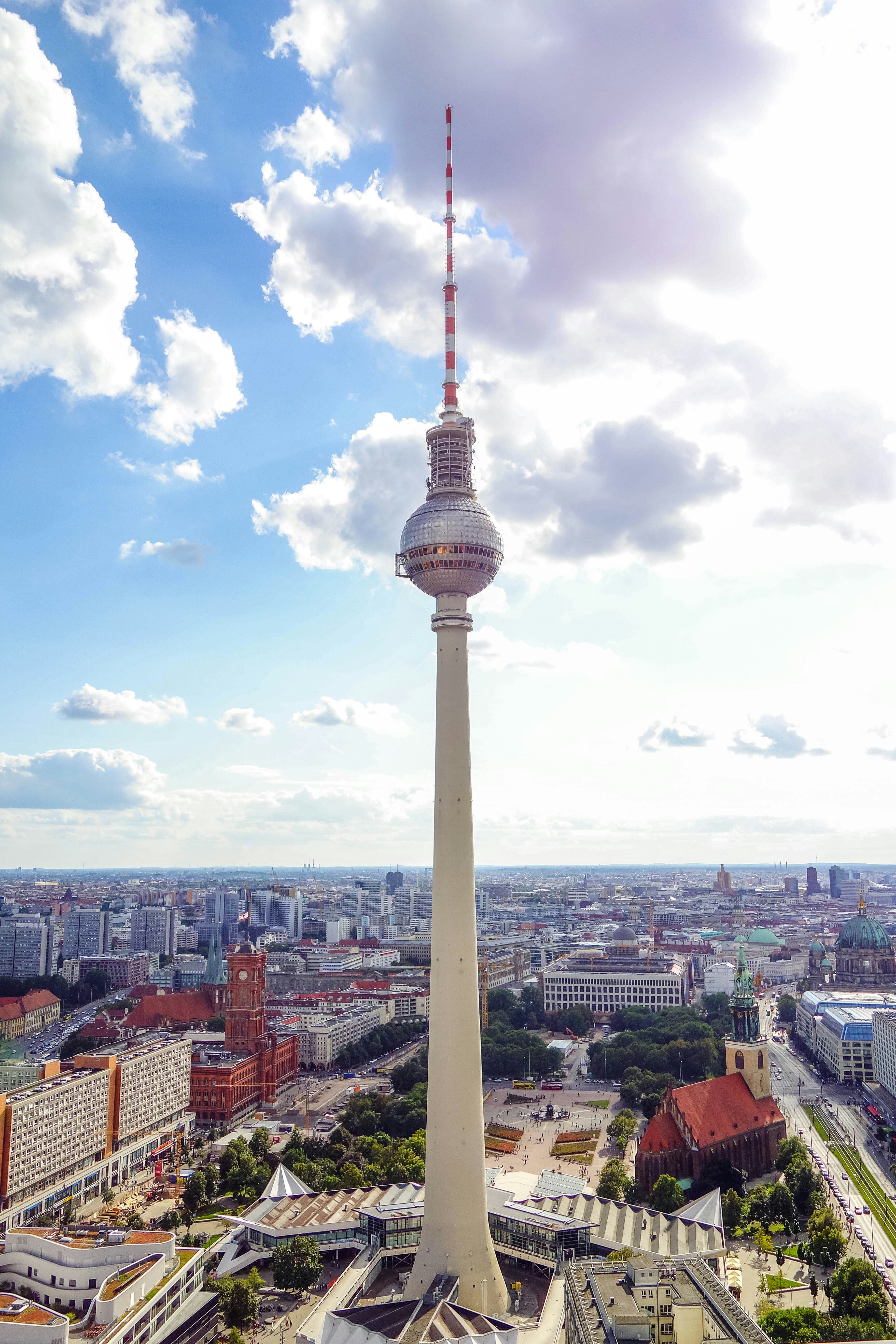 Berlin TV Tower rising above the city skyline. The Berliner Fernsehturm stands at the center of Alexanderplatz, its steel sphere catching the afternoon light above a sprawl of rooftops. Below, the red brick of the Rotes Rathaus and the green dome of Berlin Cathedral anchor the historic core, while cranes mark the city's constant rebuilding. At 368 metres it remains the tallest structure in Germany and the clearest landmark on the Berlin skyline, visible from nearly every district.