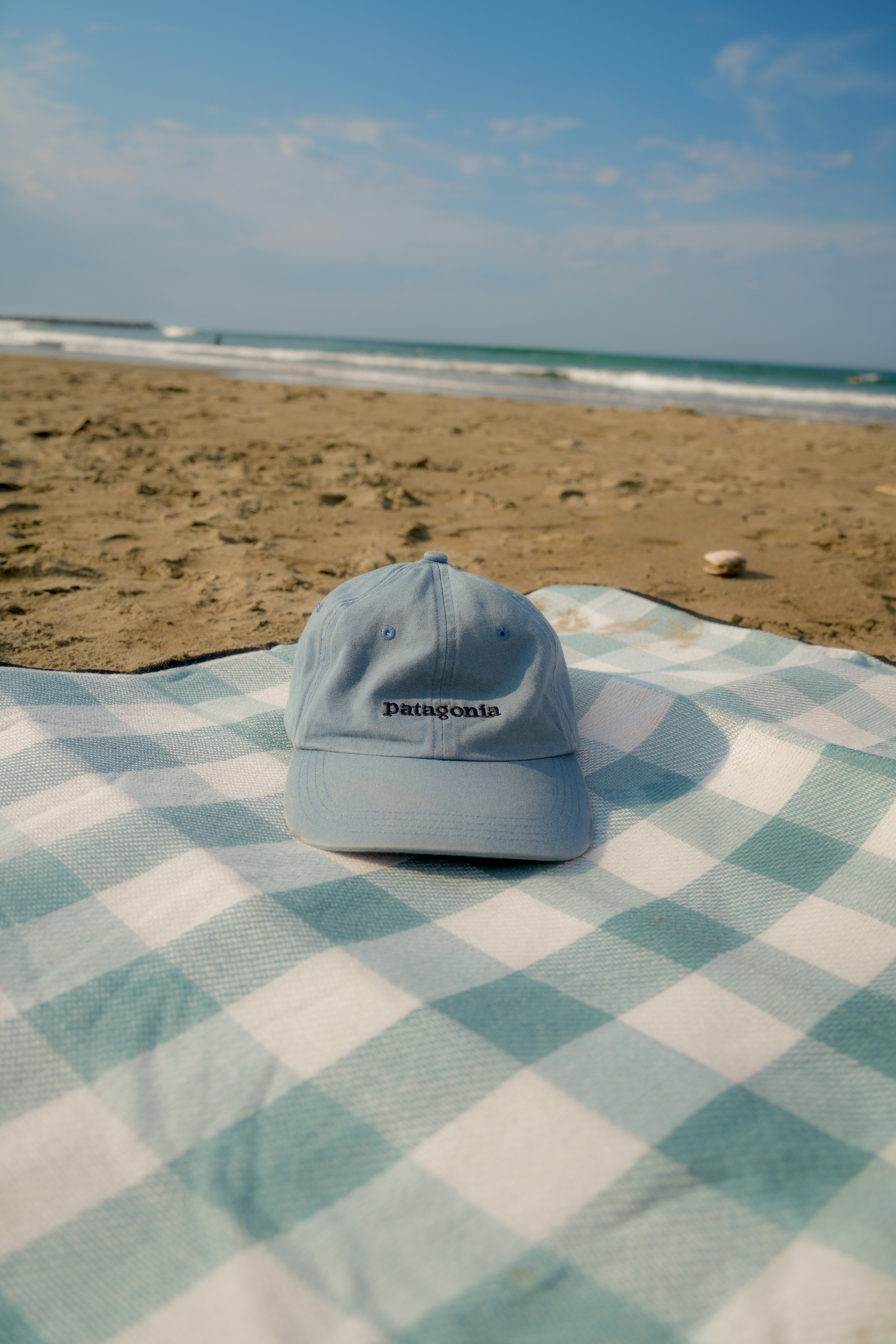 A light blue Patagonia cap rests on a blue-and-white checkered blanket spread over sandy beach with the ocean and clear sky in the background. The scene emphasizes a relaxed, sunlit beachfront moment.