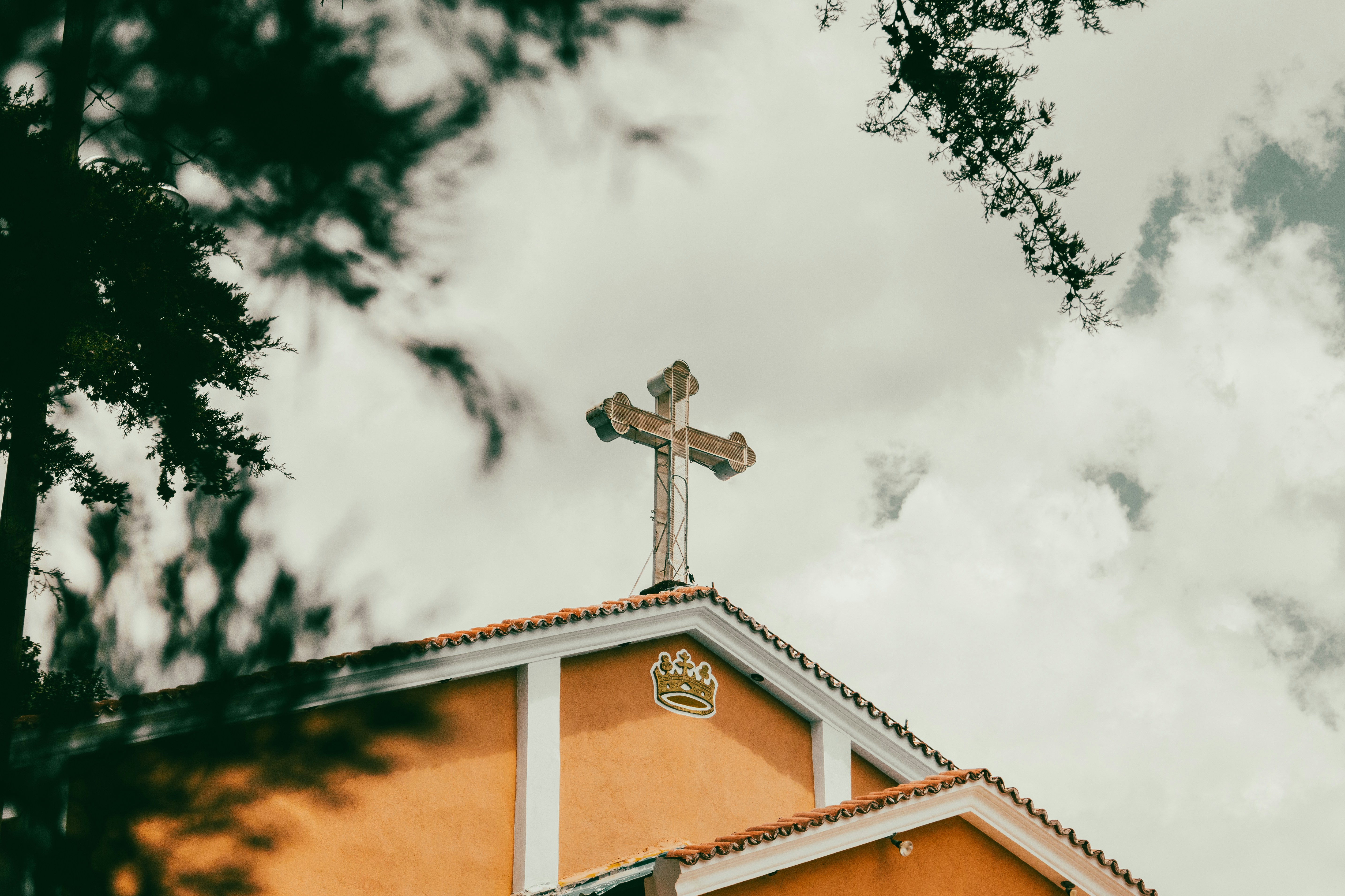 Warm-toned chapel peak with cross rises against a cloudy sky; foreground silhouettes of trees frame the scene.