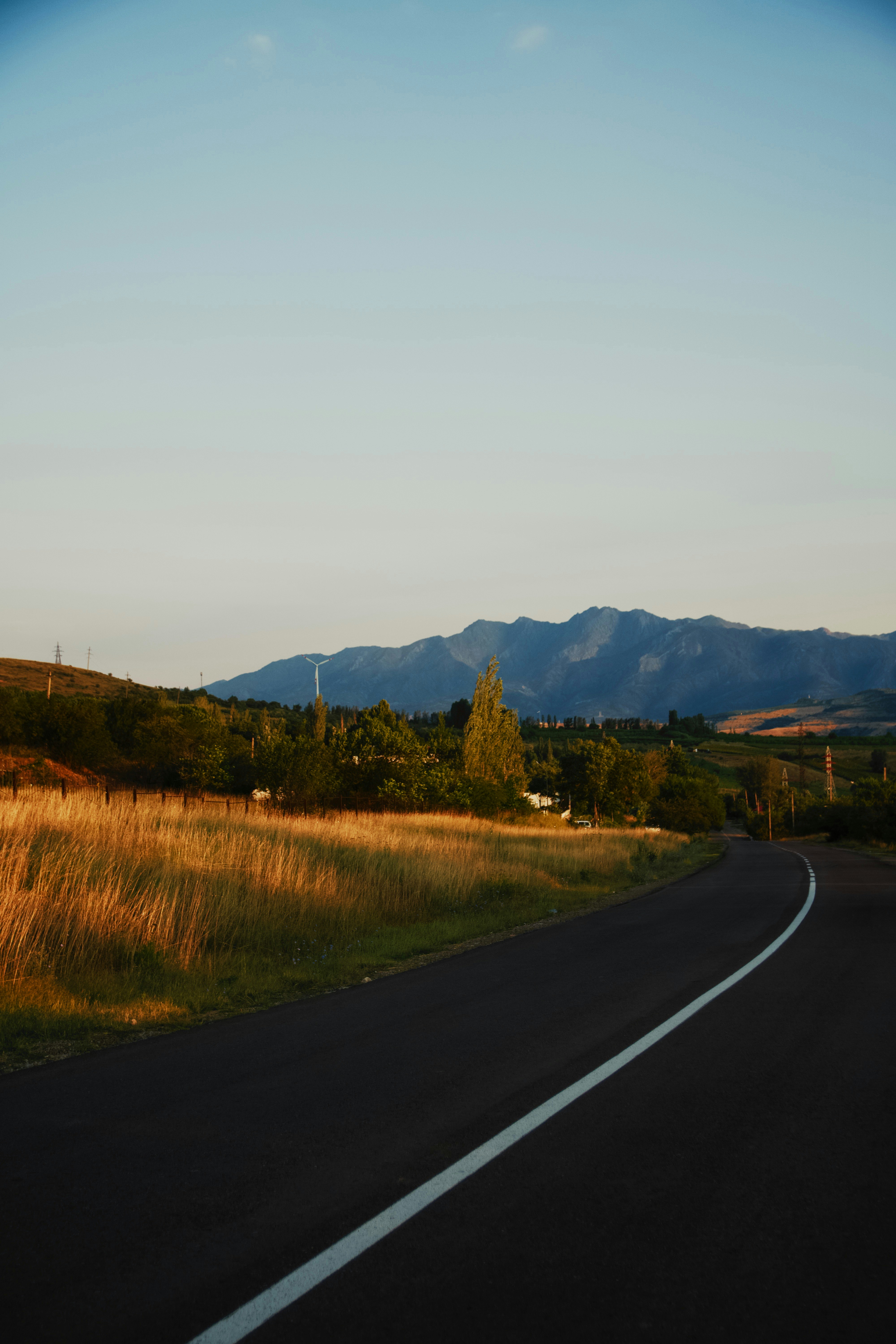 Golden-hour road winds toward a distant mountain ridge with wind turbines and scattered trees, bathed in warm light. The scene emphasizes open landscape and leading lines.