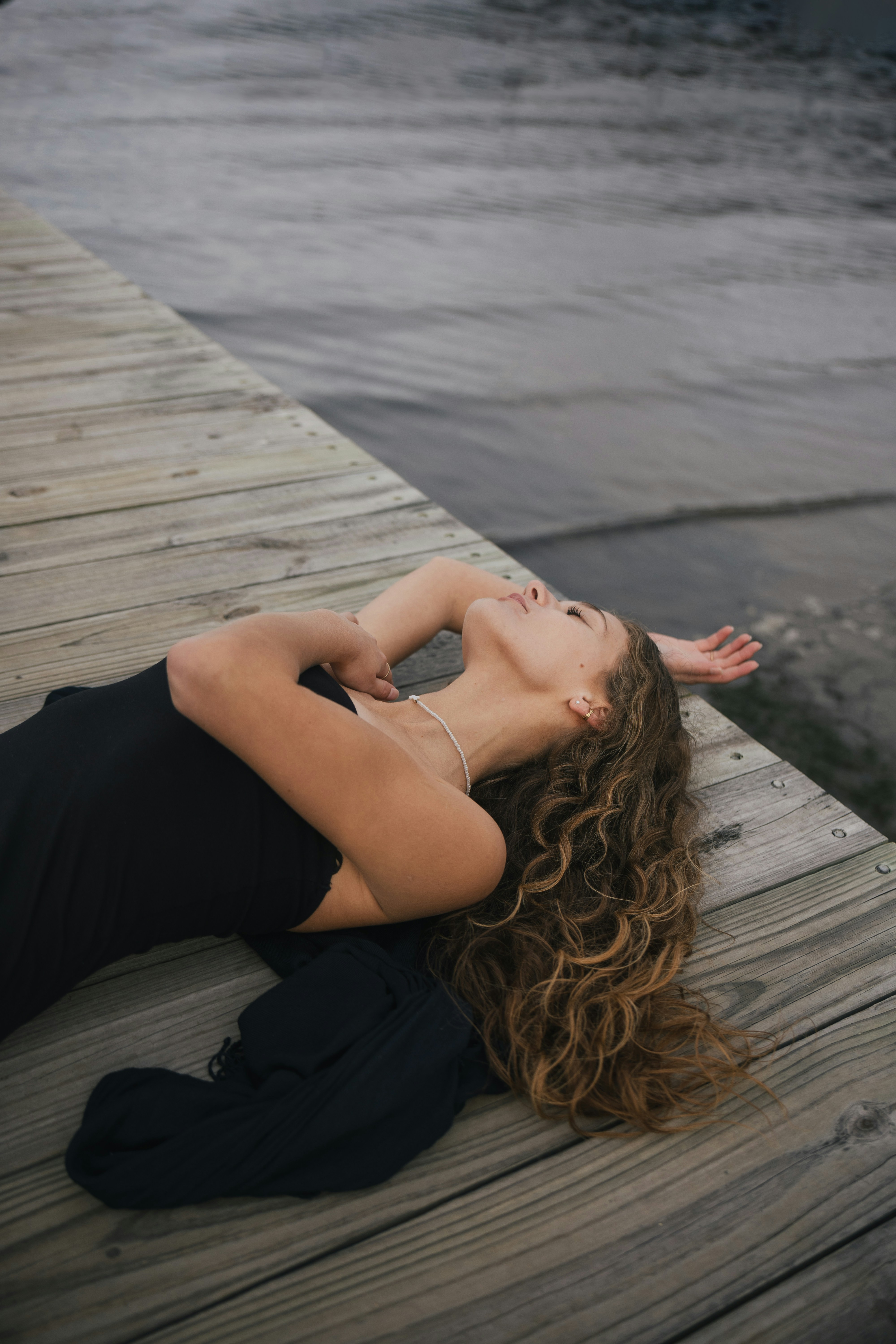 Portrait of a woman in a black dress lying on a wooden dock by calm water, eyes closed and arms raised above her head. A minimalist lakeside scene captured with natural light.