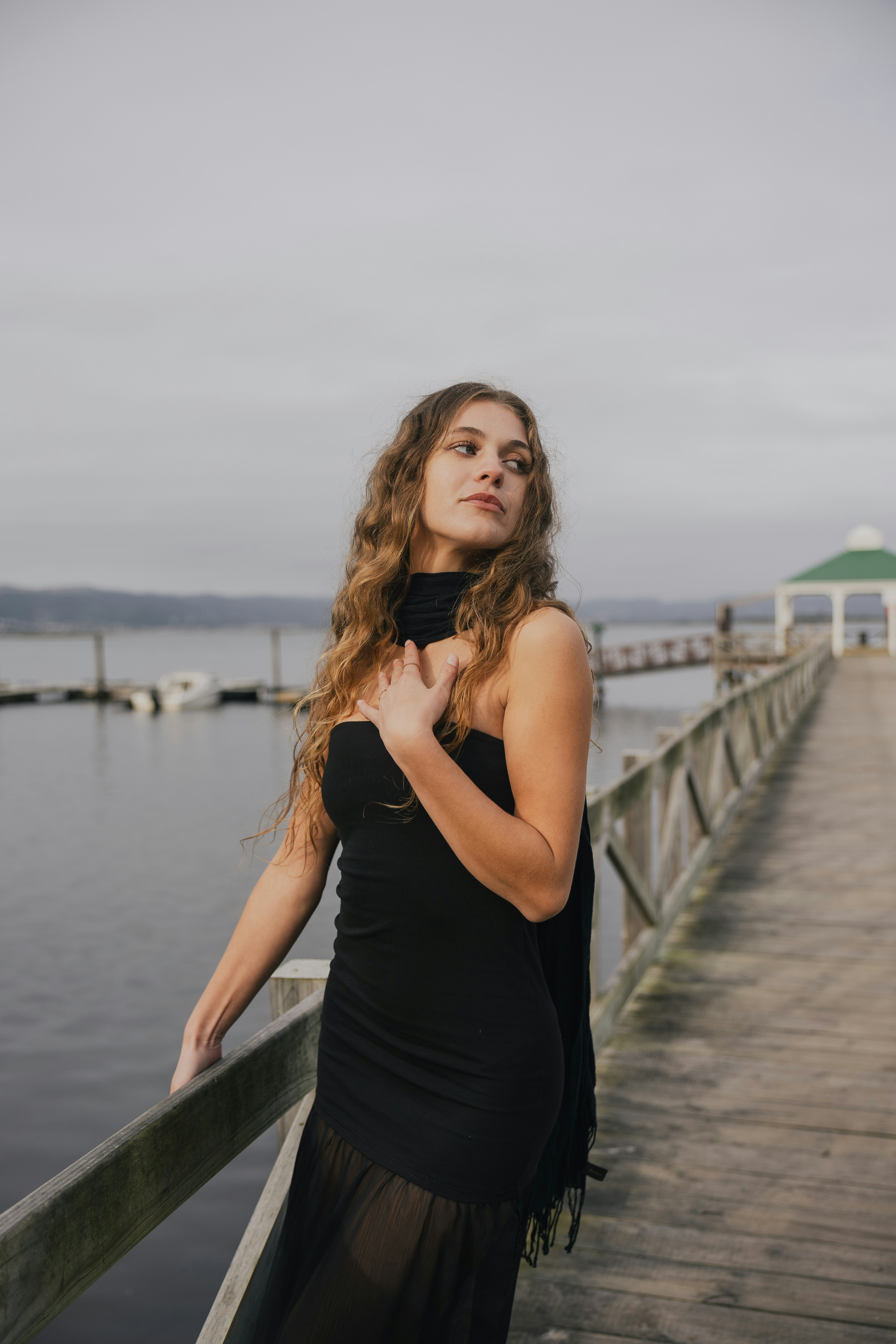 A woman in a sleek black dress leans against a railing by a calm dock, with a distant shoreline and pale sky in the background. This is a portrait-style scene captured at the waterfront.
