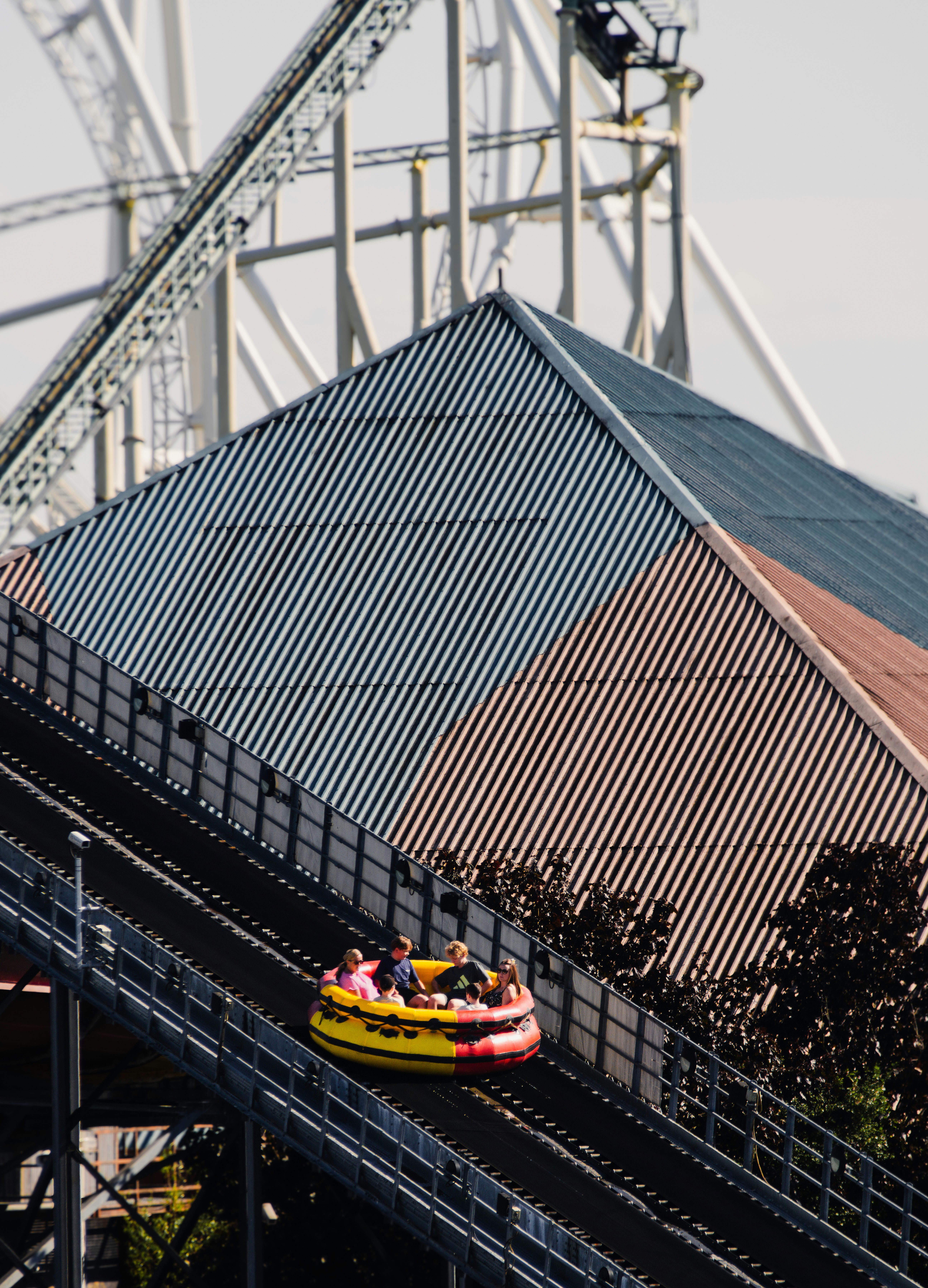 Group of riders in a yellow circular raft descends a slate-gray roller coaster track beneath angular industrial roofs.