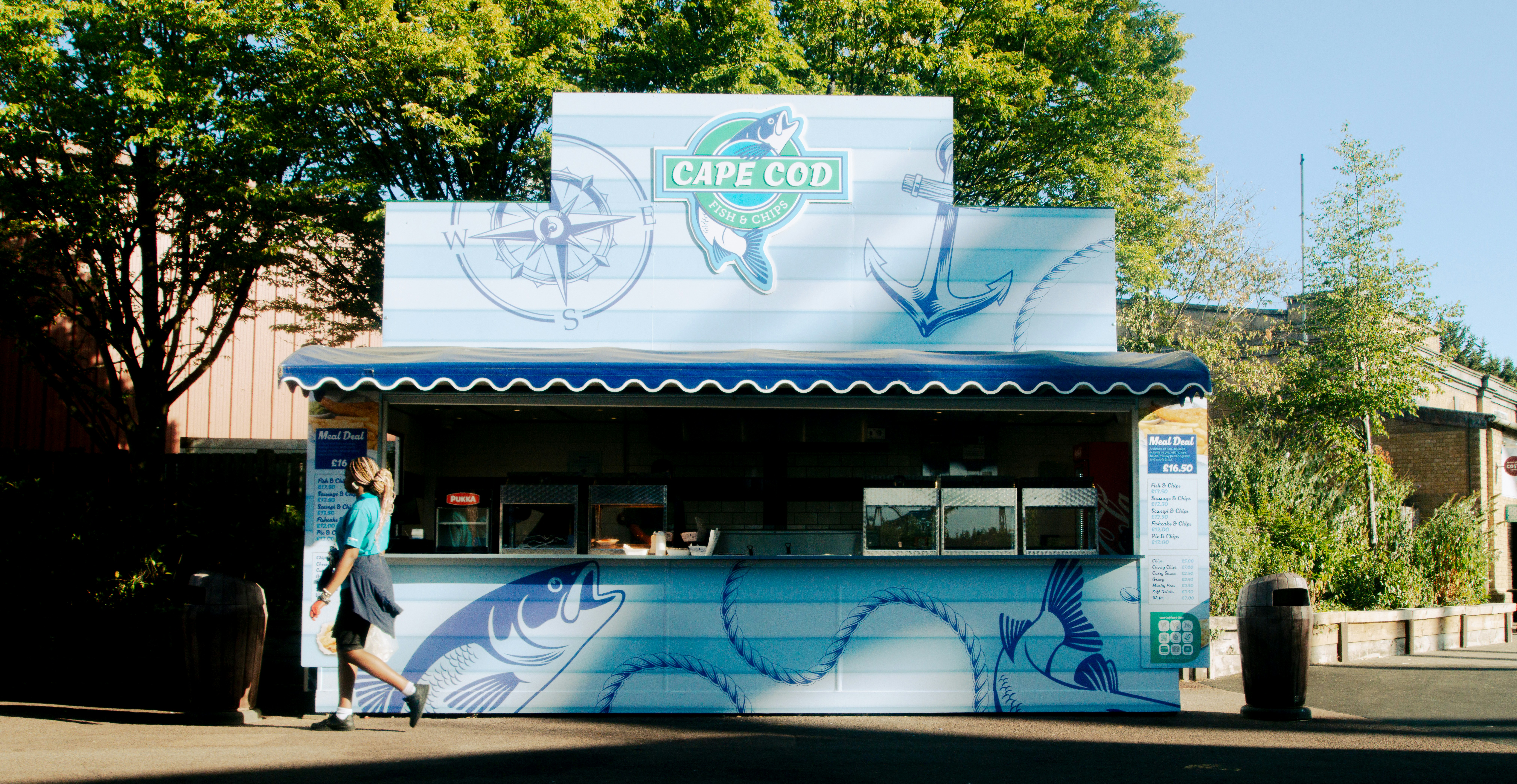 Colorful blue food shack with a wavy design and a sign Cape Cod above the service window; a person walks past on the left while trees frame the scene.