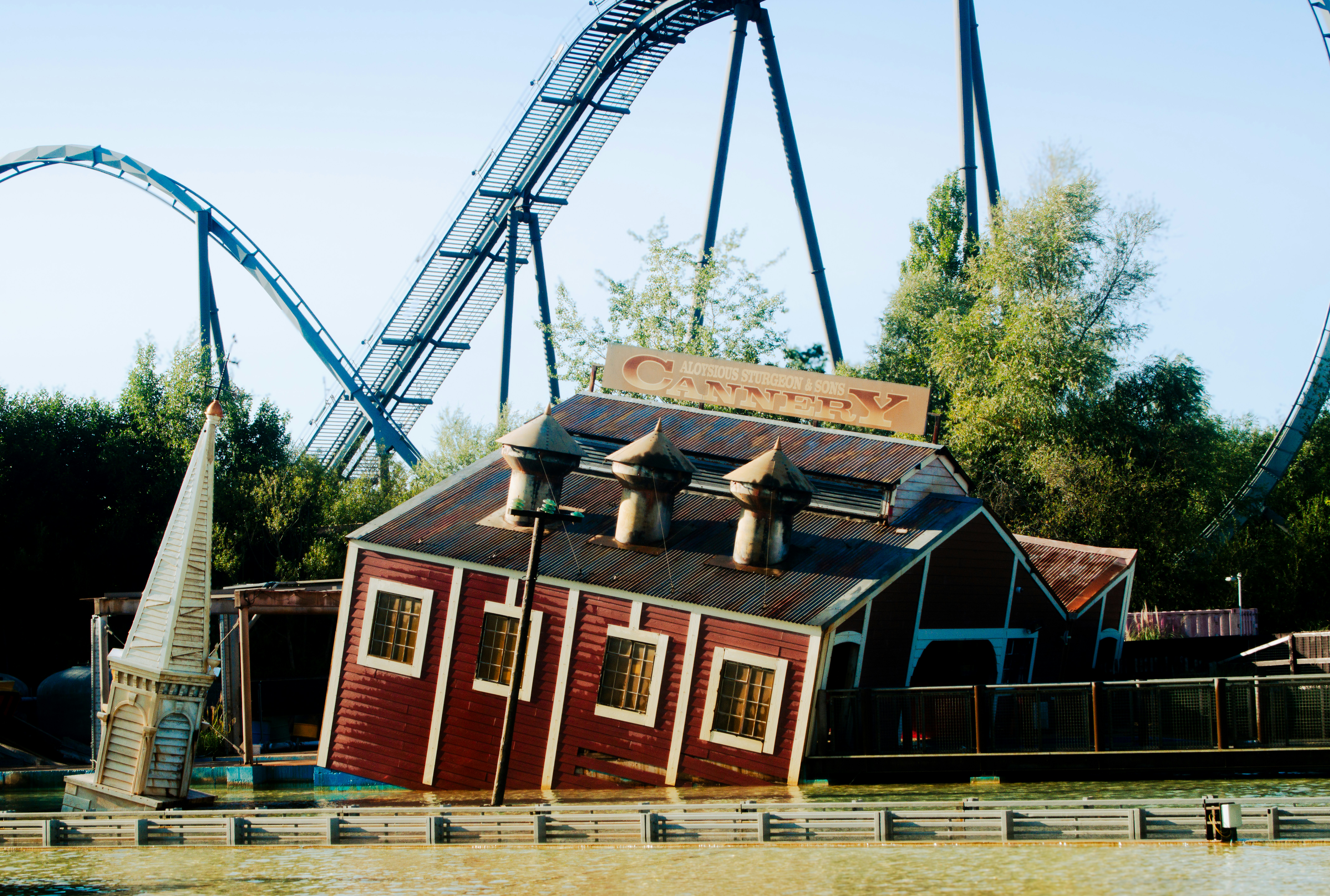 A tilted red wooden cabin sits near water with a roller coaster and blue sky in the background, suggesting an amusement park setting.