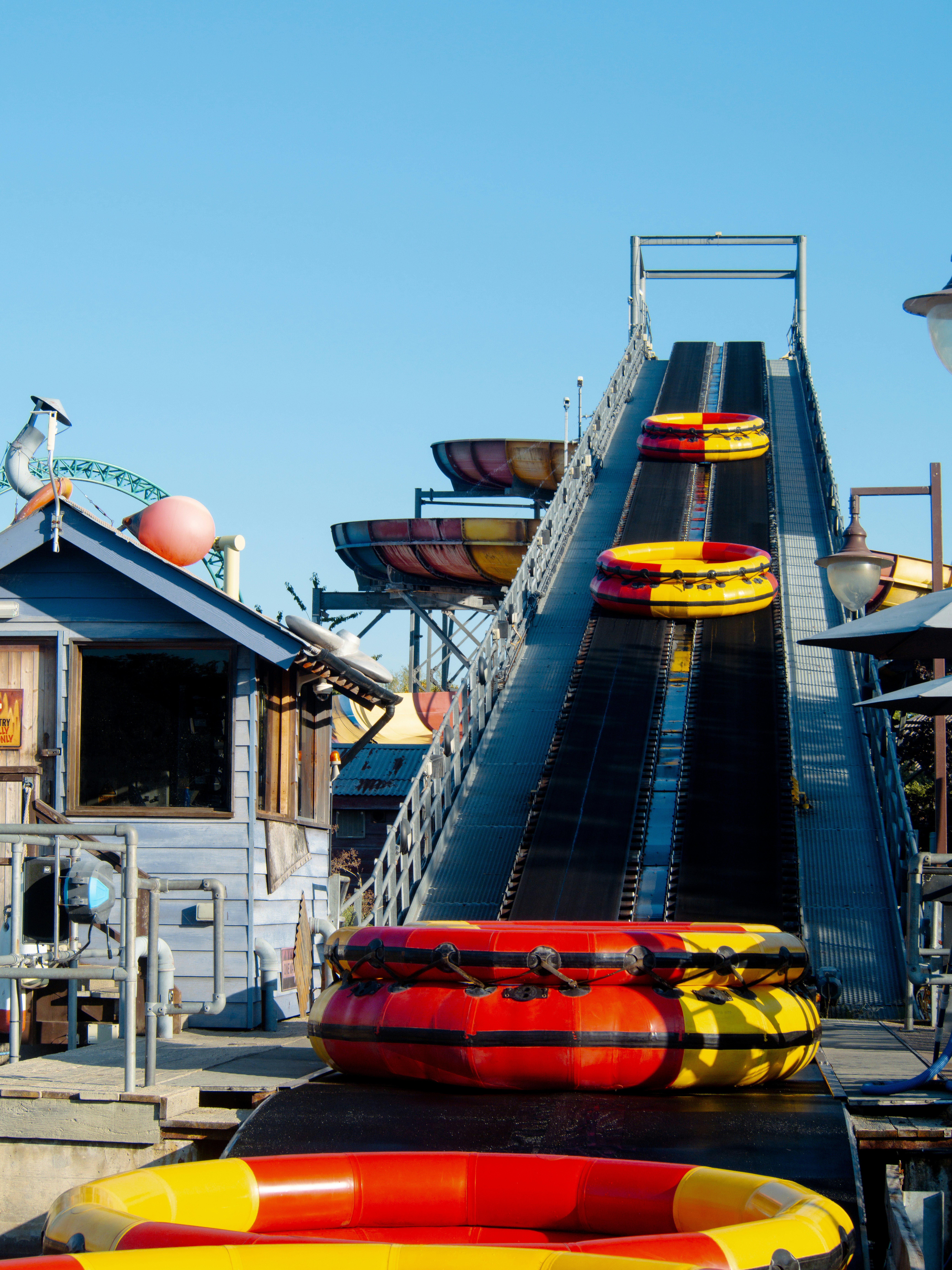 Colorful rafts line a tall slide on a pier, with a small control shack to the left and a clear blue sky above.