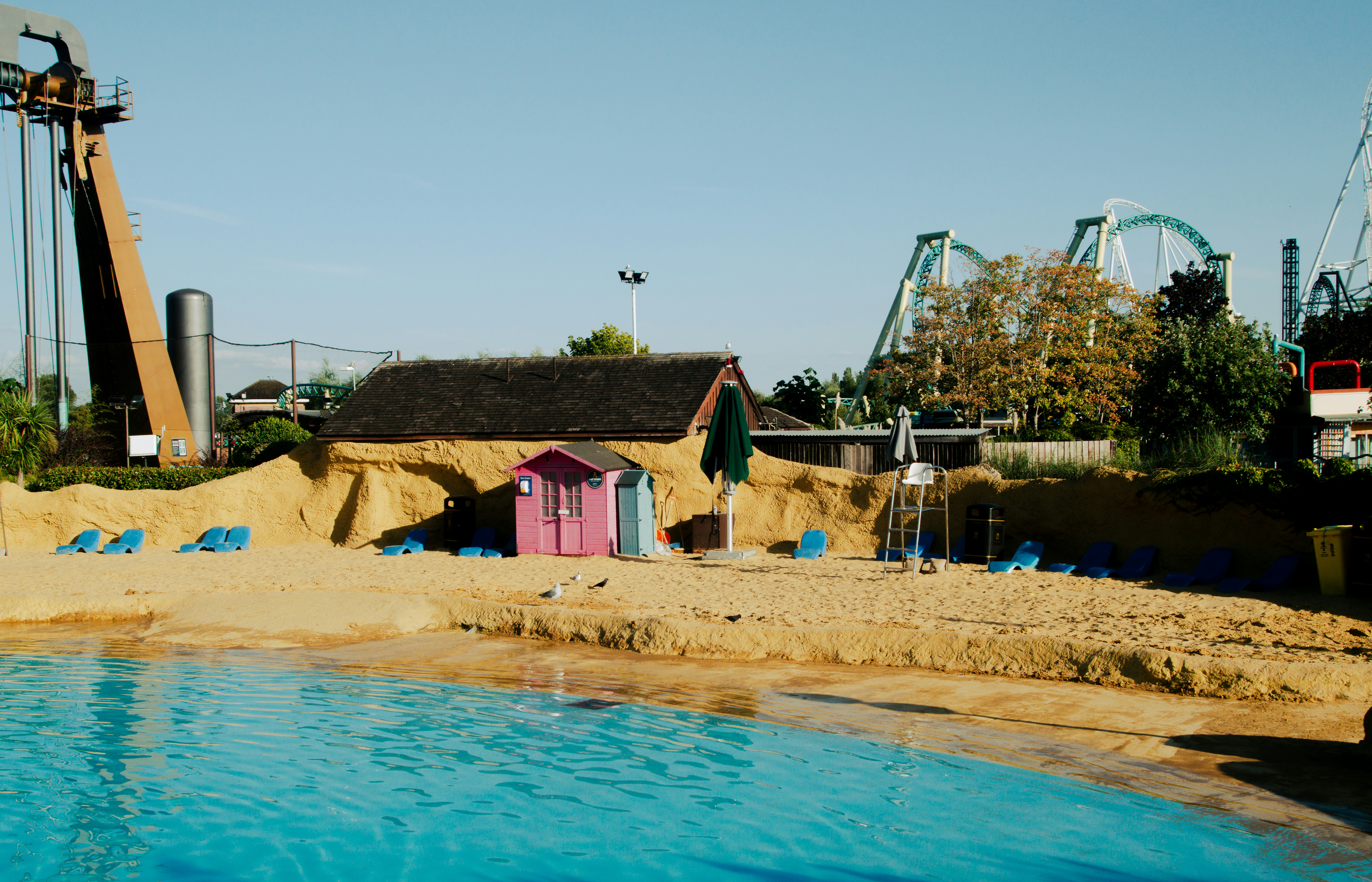 Bright pool-edge scene with a sandy shore, a small pink hut, and blue lounge chairs under a clear sky.