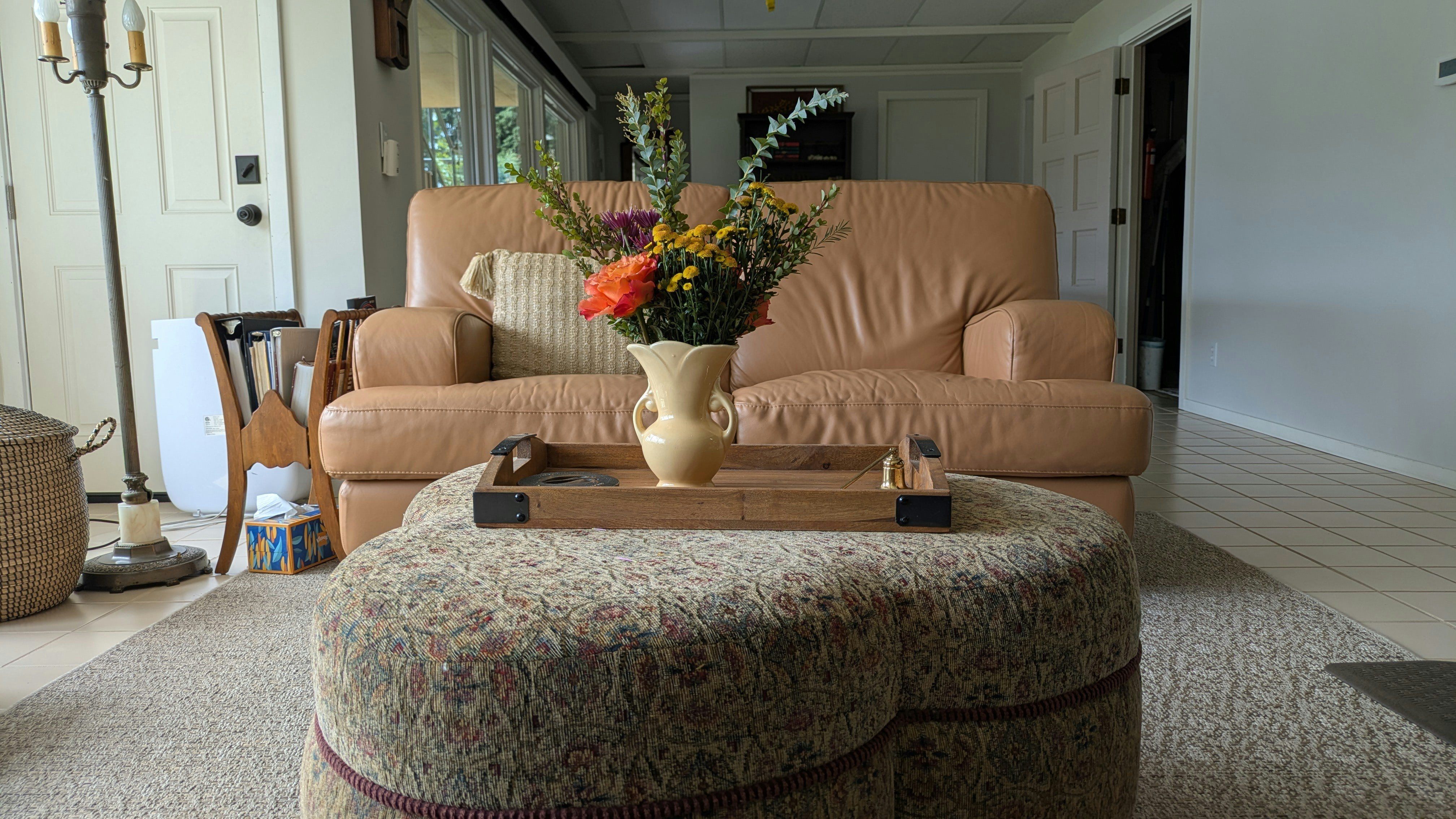 A bunch of assorted flowers in a cream colored vase with a caramel toned leather couch behind it.