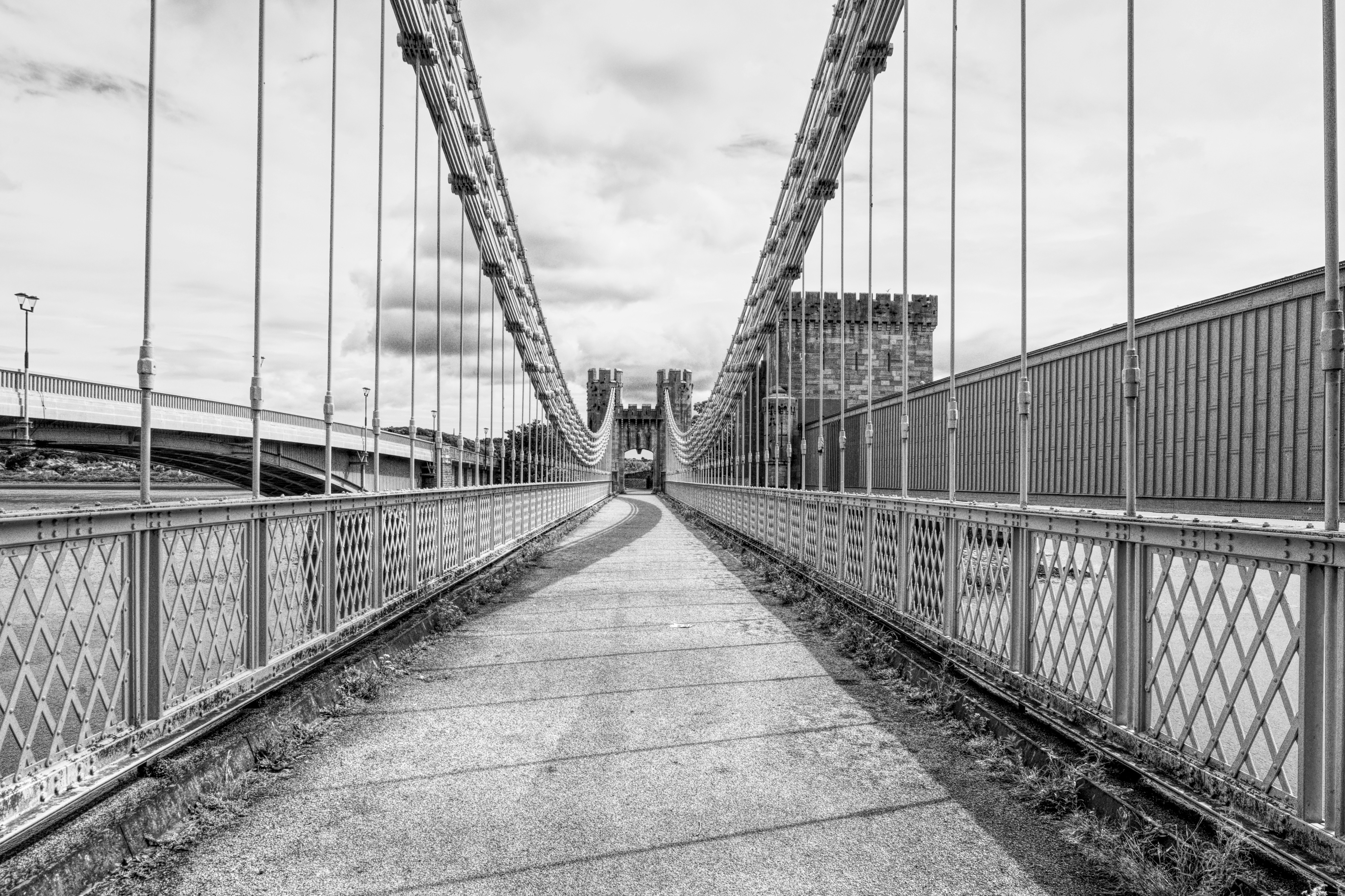 Monochrome view of a long suspension bridge walkway receding toward a distant tower, emphasizing converging lines and perspective in a stark urban landscape.