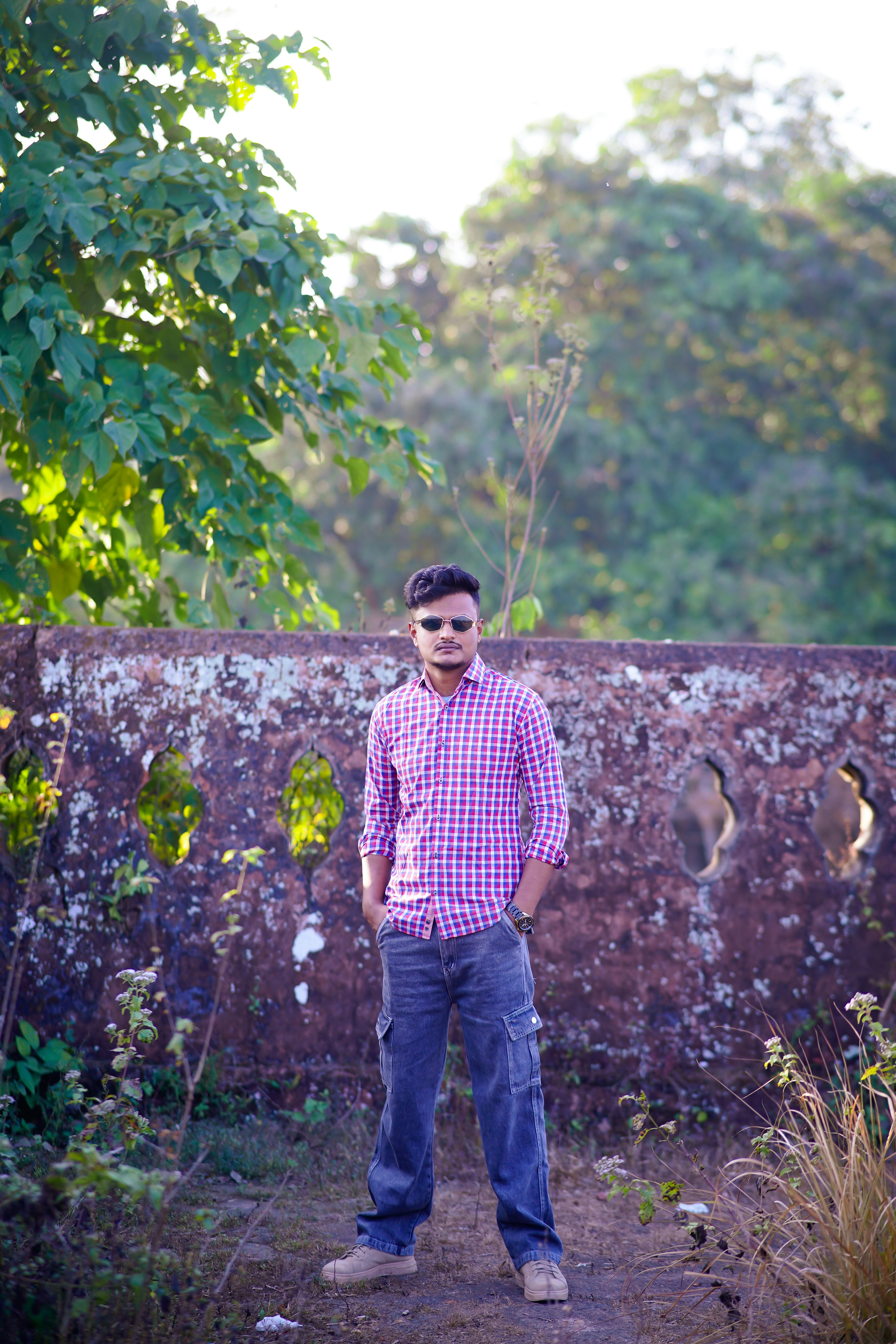 Man in Checkered Shirt Stands Against Weathered Wall with Greenery Backdrop