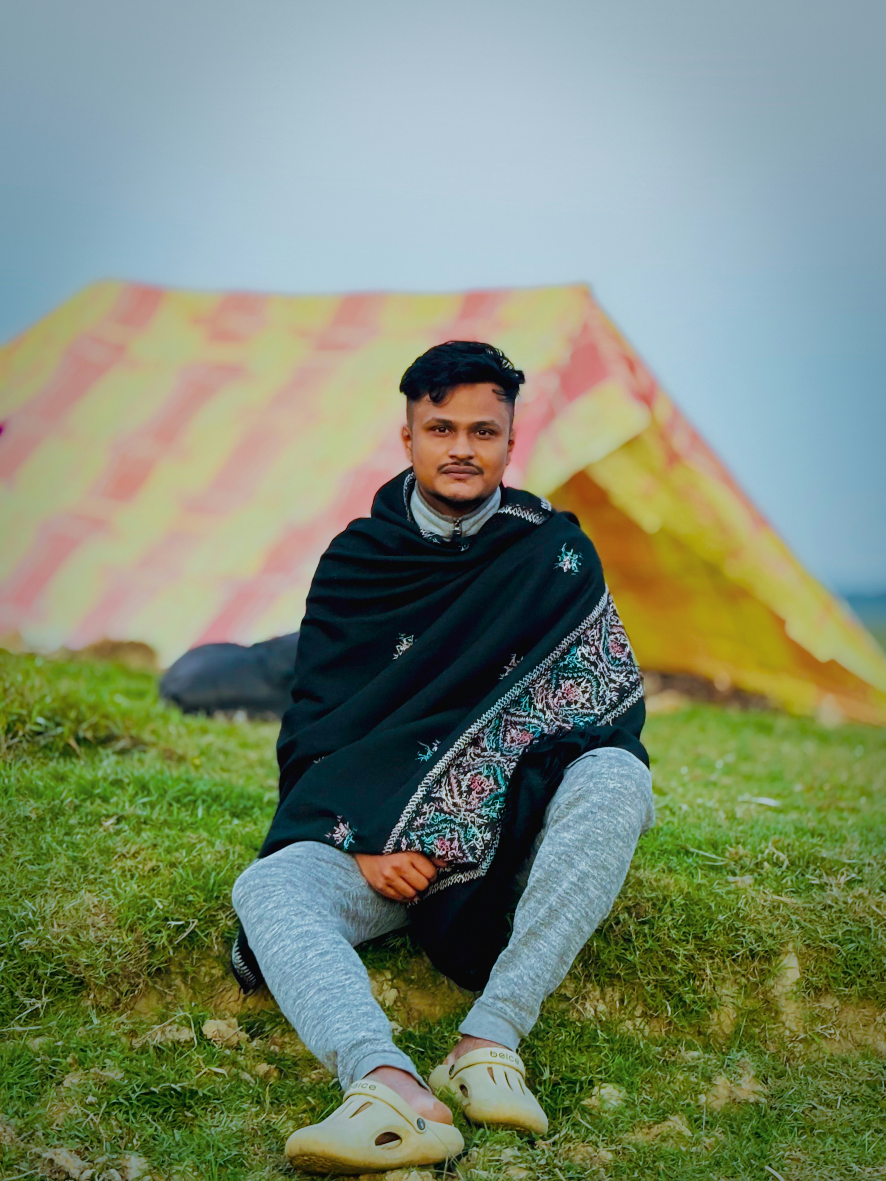 Traveler in a patterned shawl rests before a striped shelter on green hillside