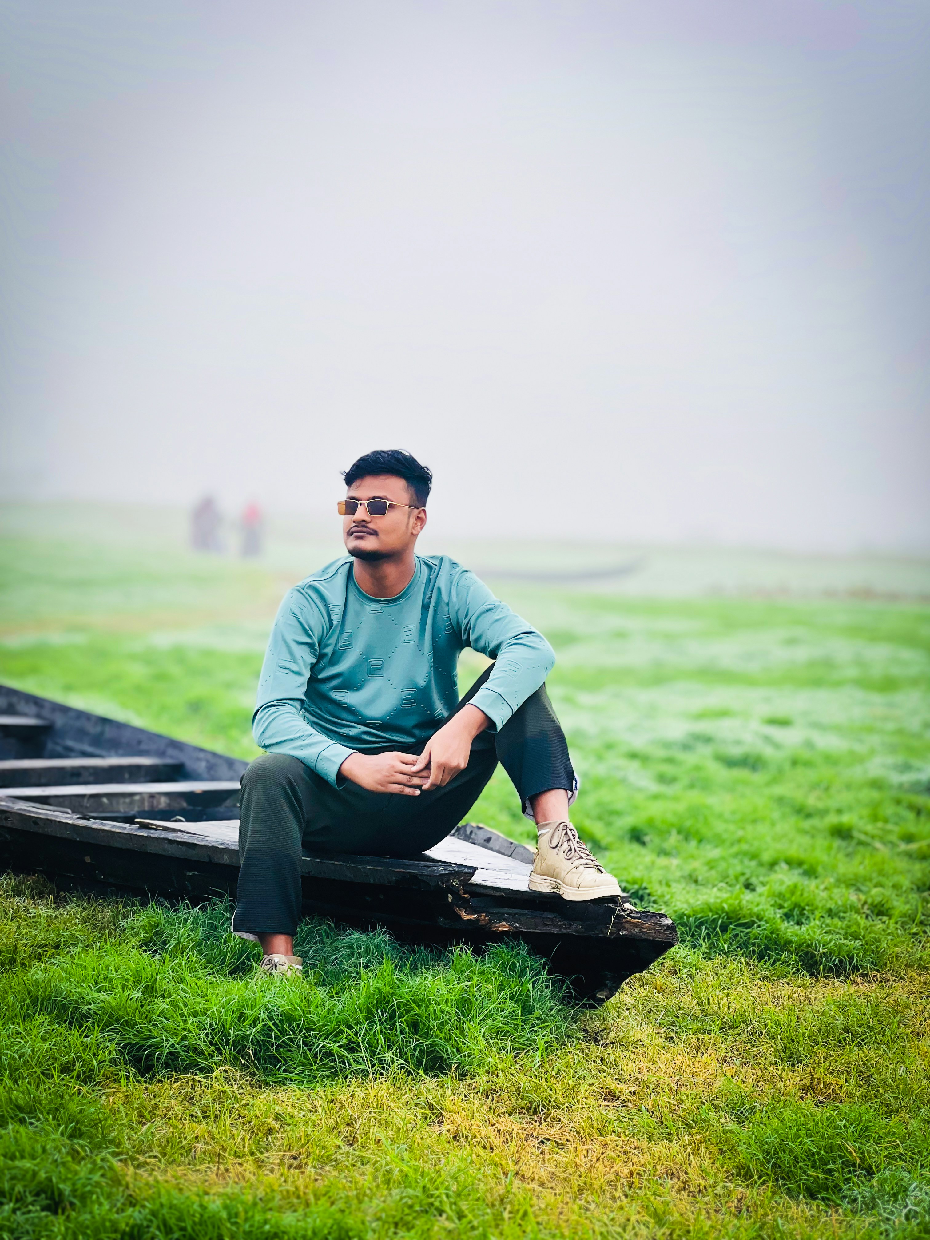 Man in teal sweater sits contemplatively on a weathered boat amid a foggy lakeside meadow
