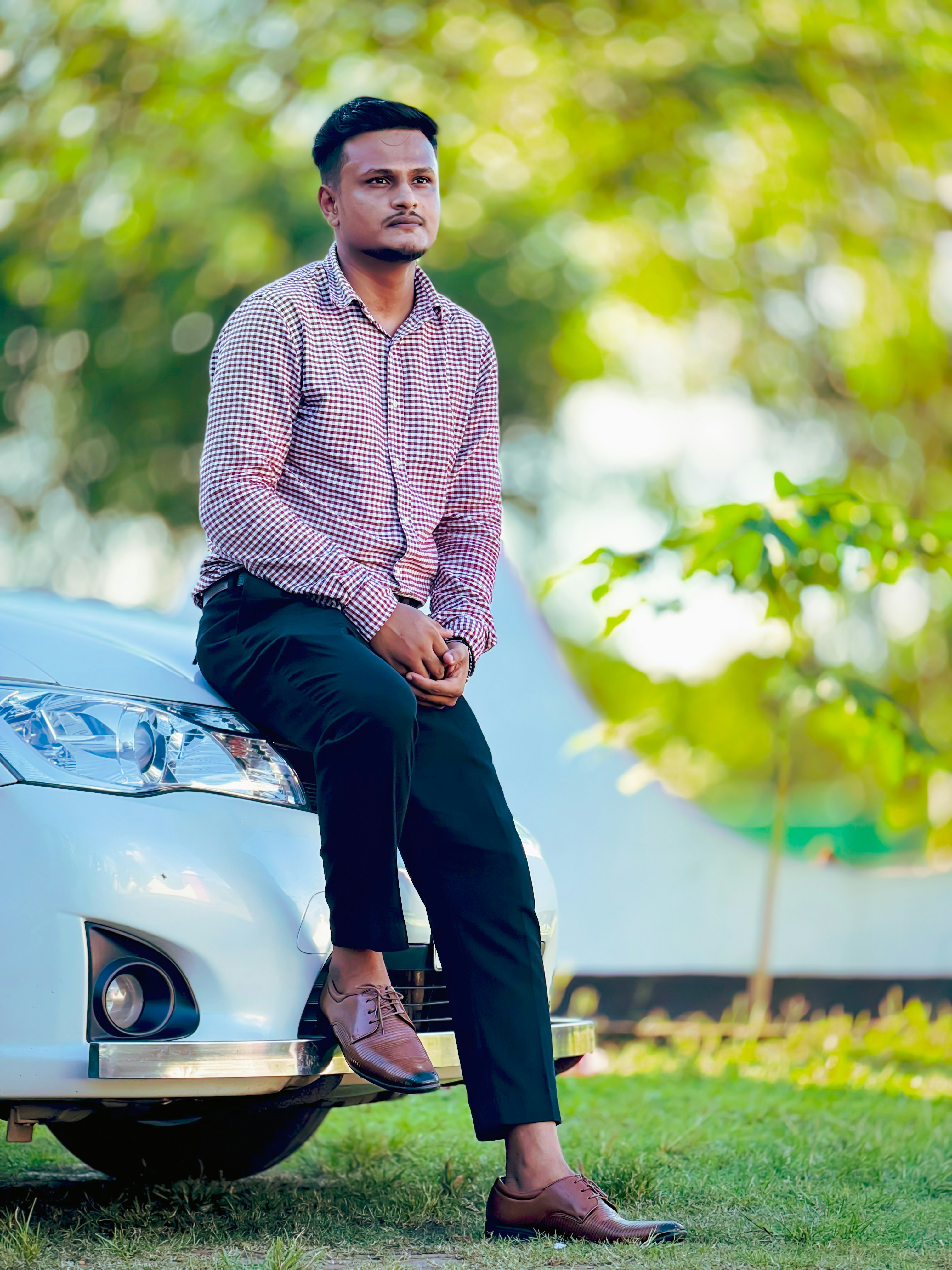 Man in checked shirt resting on car hood amid sunlit greenery